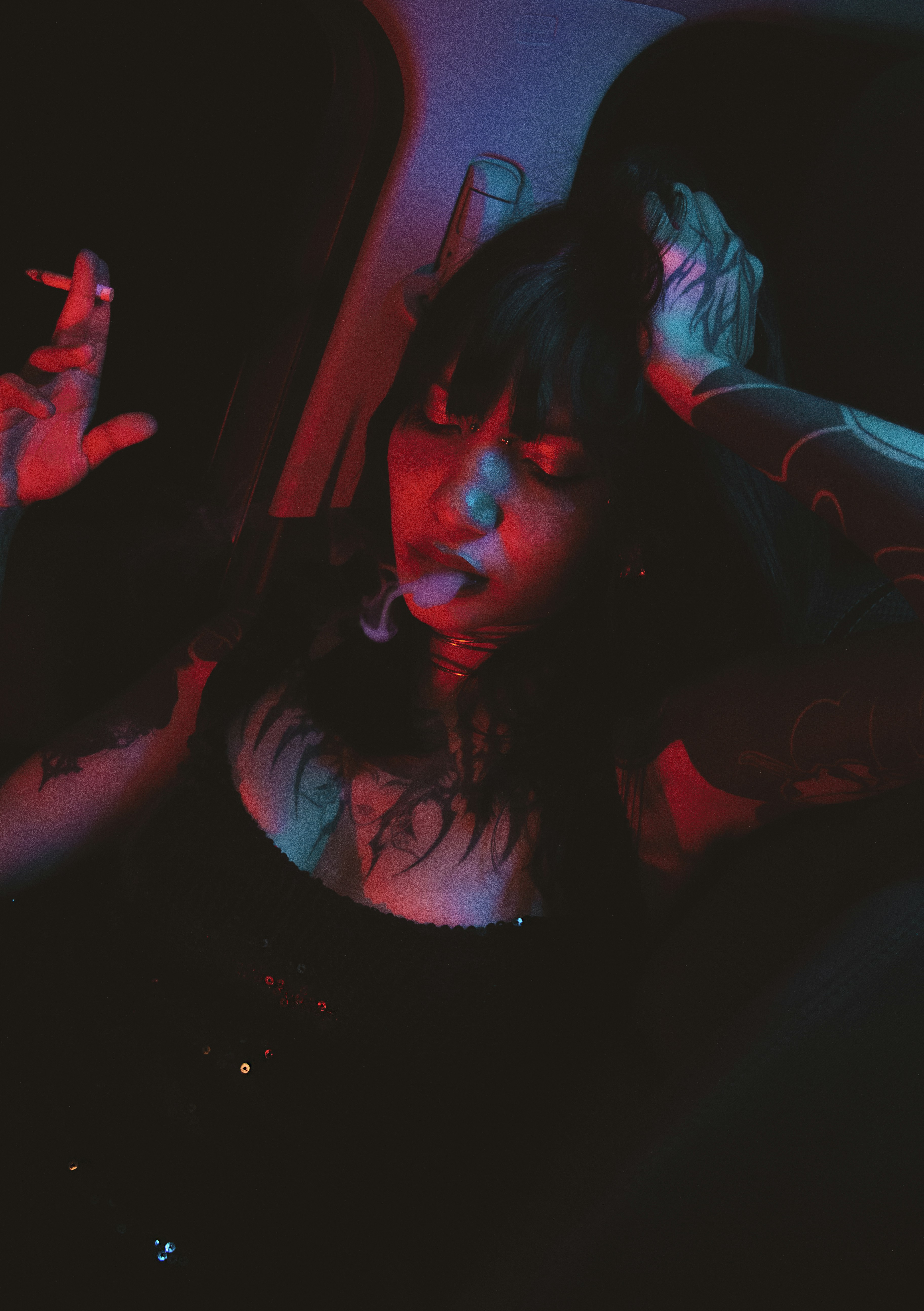A figure exudes a moody ambiance, enveloped in deep reds and blues, with smoke curling around them, suggesting a moment of introspection. The contrast highlights intricate tattoos and a reflective expression.