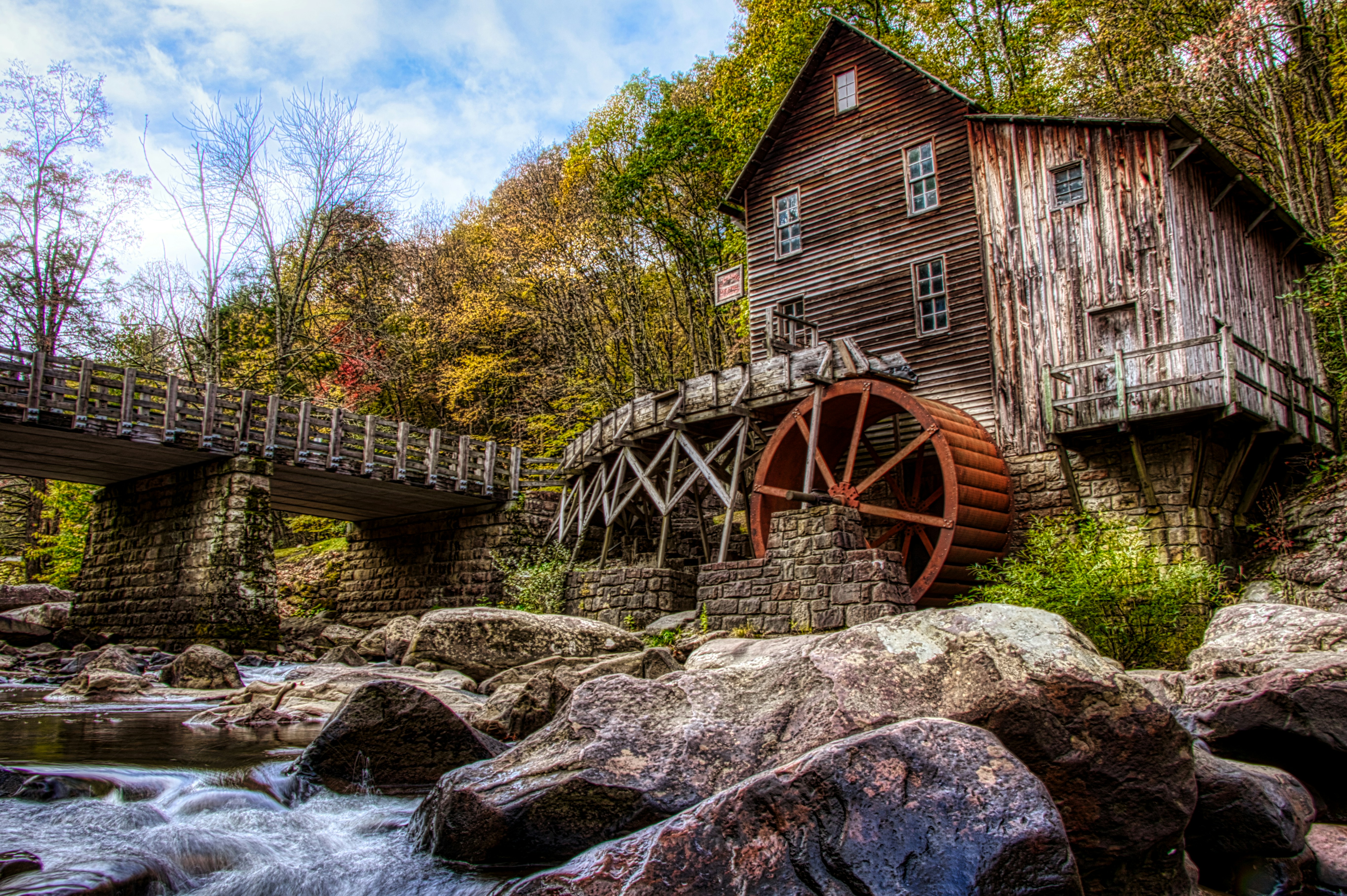 Historic watermill nestled by a rocky stream, surrounded by autumn foliage and a rustic bridge. The waterwheel stands prominently, showcasing the charm of bygone craftsmanship.