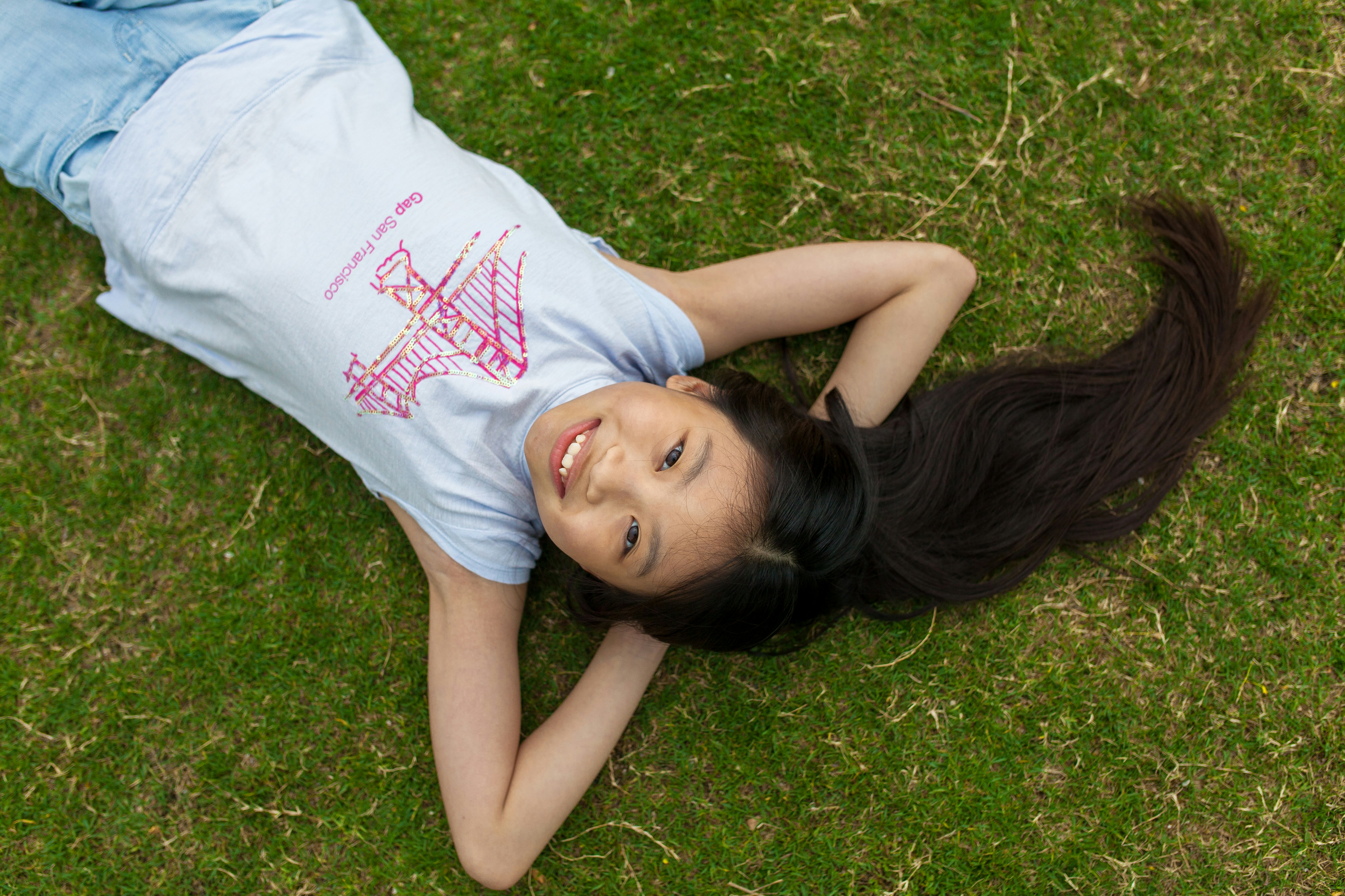 A young girl relaxes on lush grass, her long hair cascading around her as she smiles up at the sky.