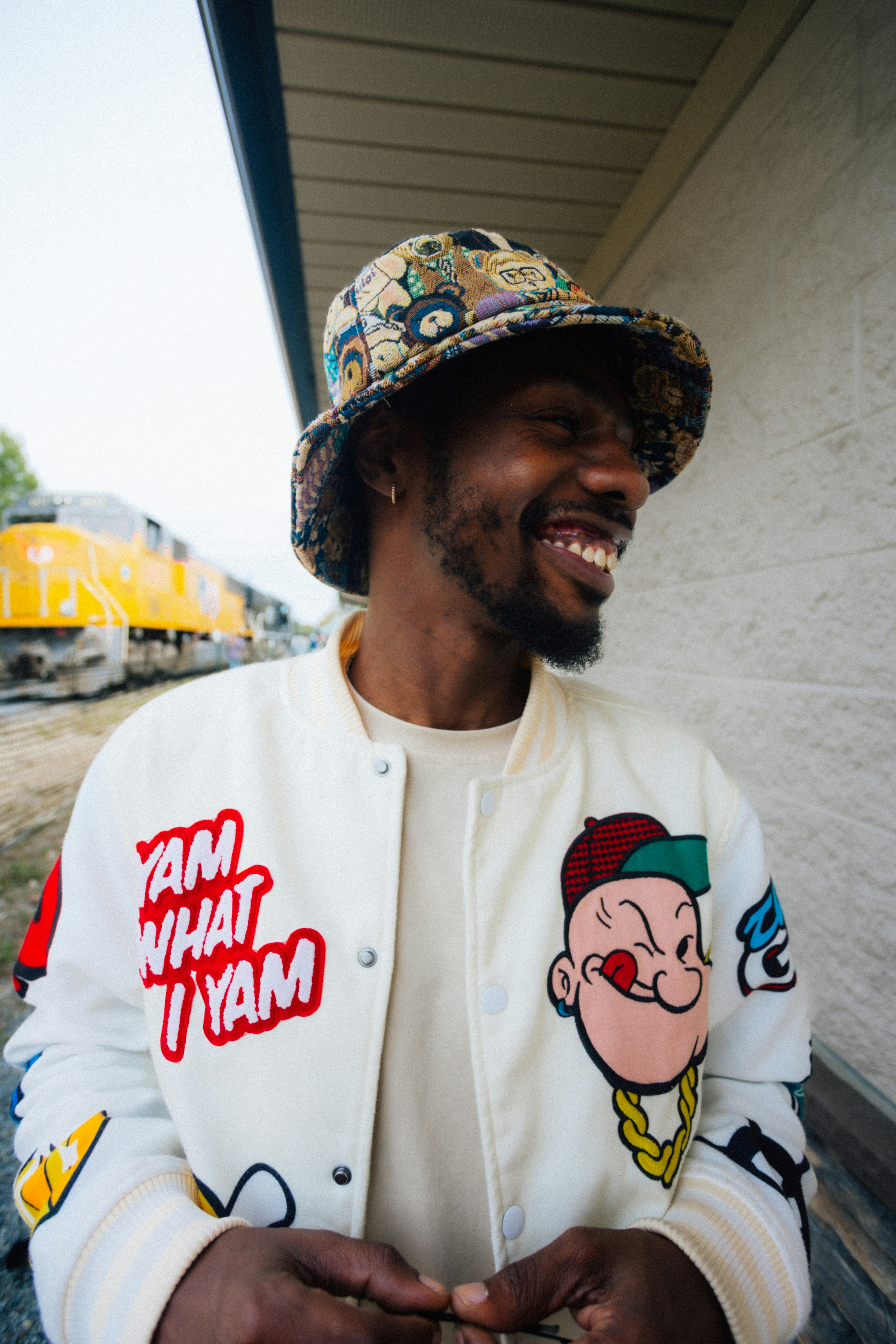 Man wearing a colorful, embroidered jacket and patterned bucket hat smiles against an urban backdrop with a train. His playful attire adds to the lively atmosphere.