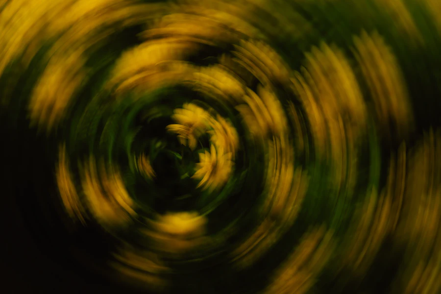 Circular motion blur of yellow flowers and green foliage spiraling around a dark center. The frame feels abstract and energetic, with the brightest details clustered near the middle.