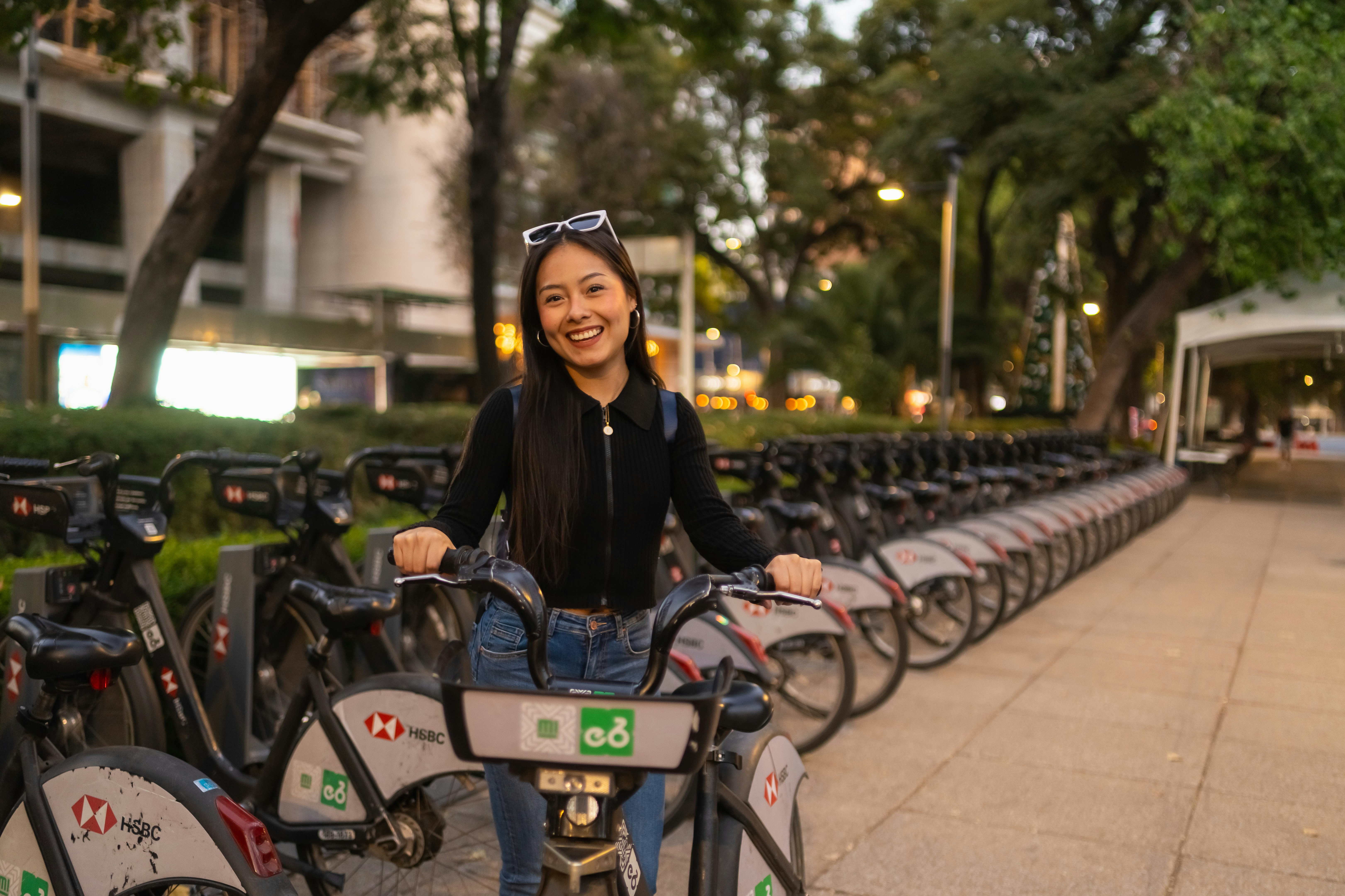 Young woman smiling while holding a bike at a bike-sharing station in an urban setting. The scene captures the essence of city life and sustainable transport.