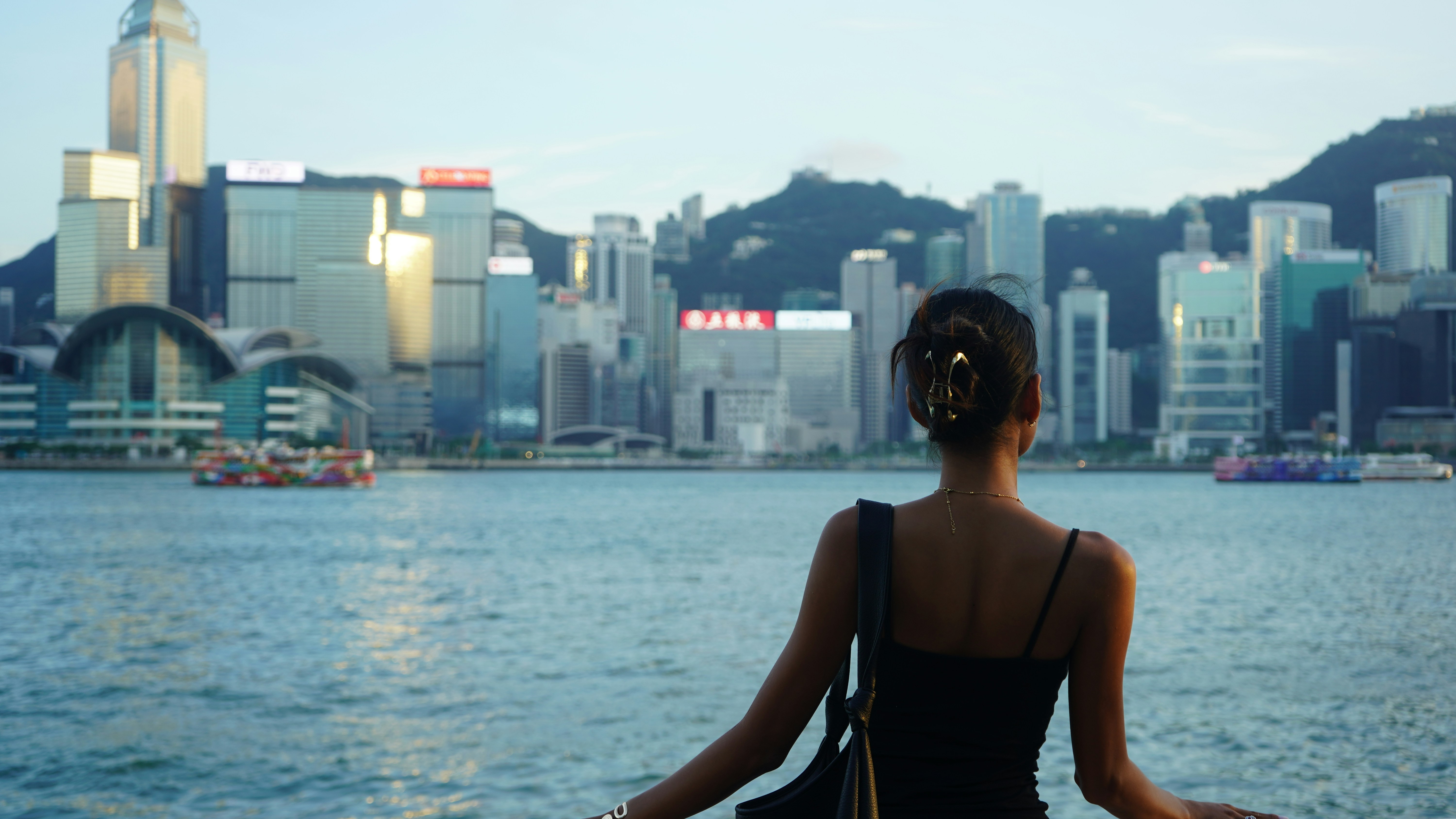 Woman gazing across Hong Kong harbour during dusk | Woman looking at hong kong skyline from across water