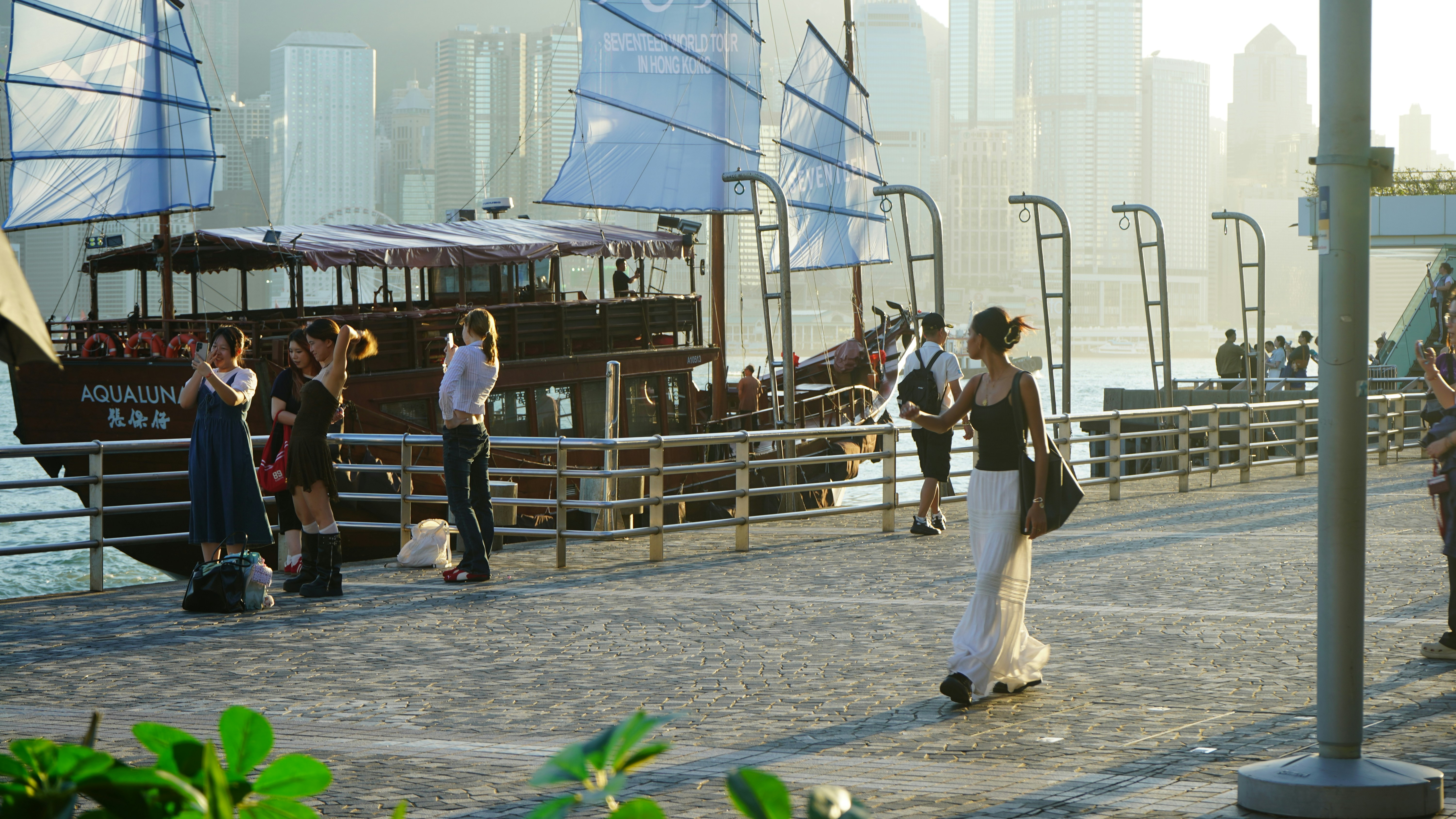Tourists and locals by the Aqualuna junk boat at golden hour | People walking on a promenade with a traditional boat.