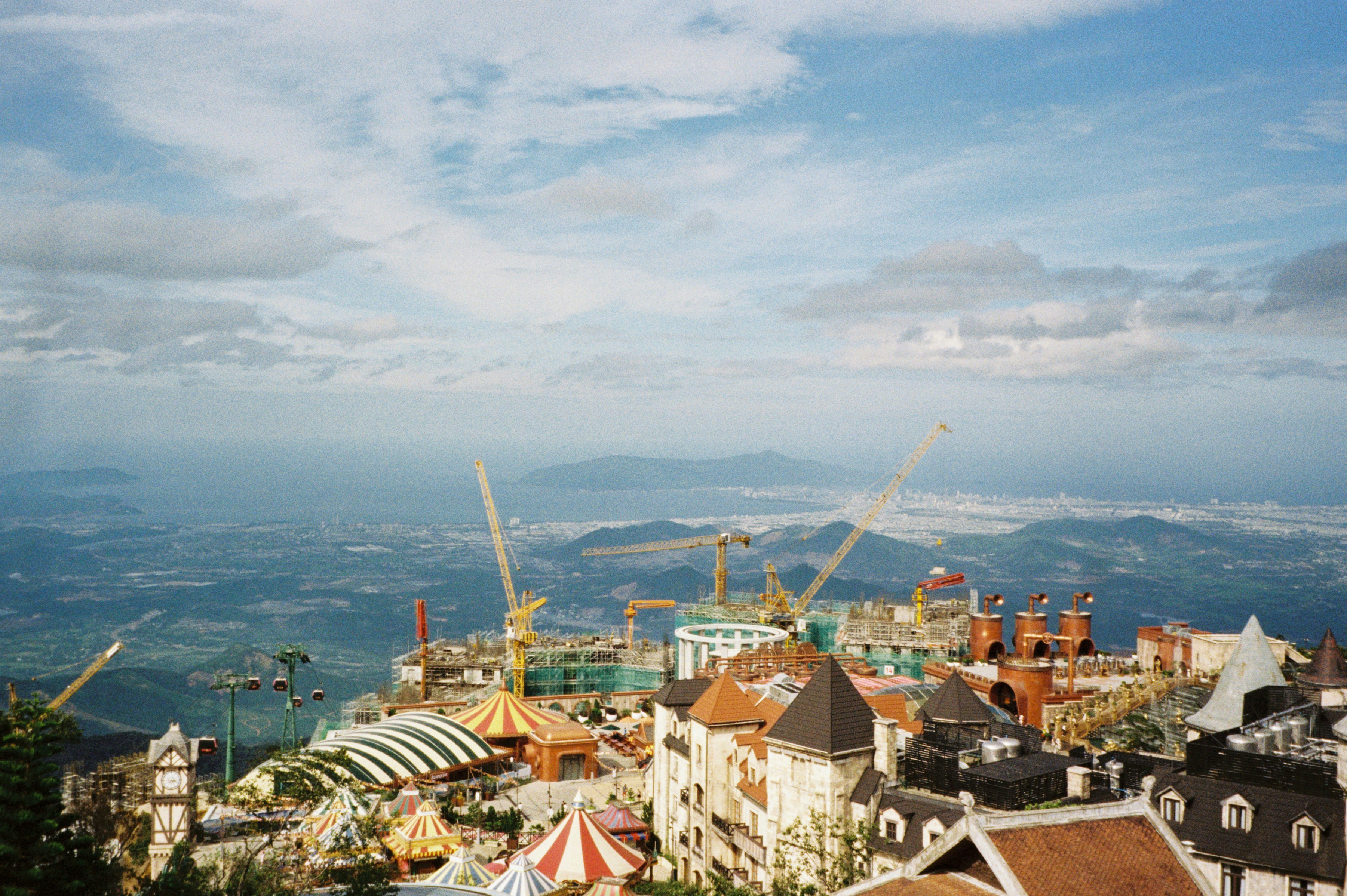 A vibrant amusement park nestled atop a mountain, surrounded by construction cranes and a distant coastal view. The scene captures the blend of leisure and development.