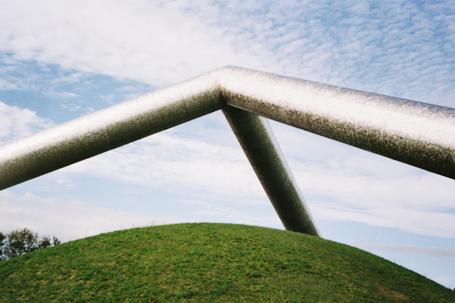 A large white metal arch structure spans over a rounded green hill beneath a pale blue, cloud-streaked sky. Mossy discoloration along the underside adds texture to the stark geometric form.