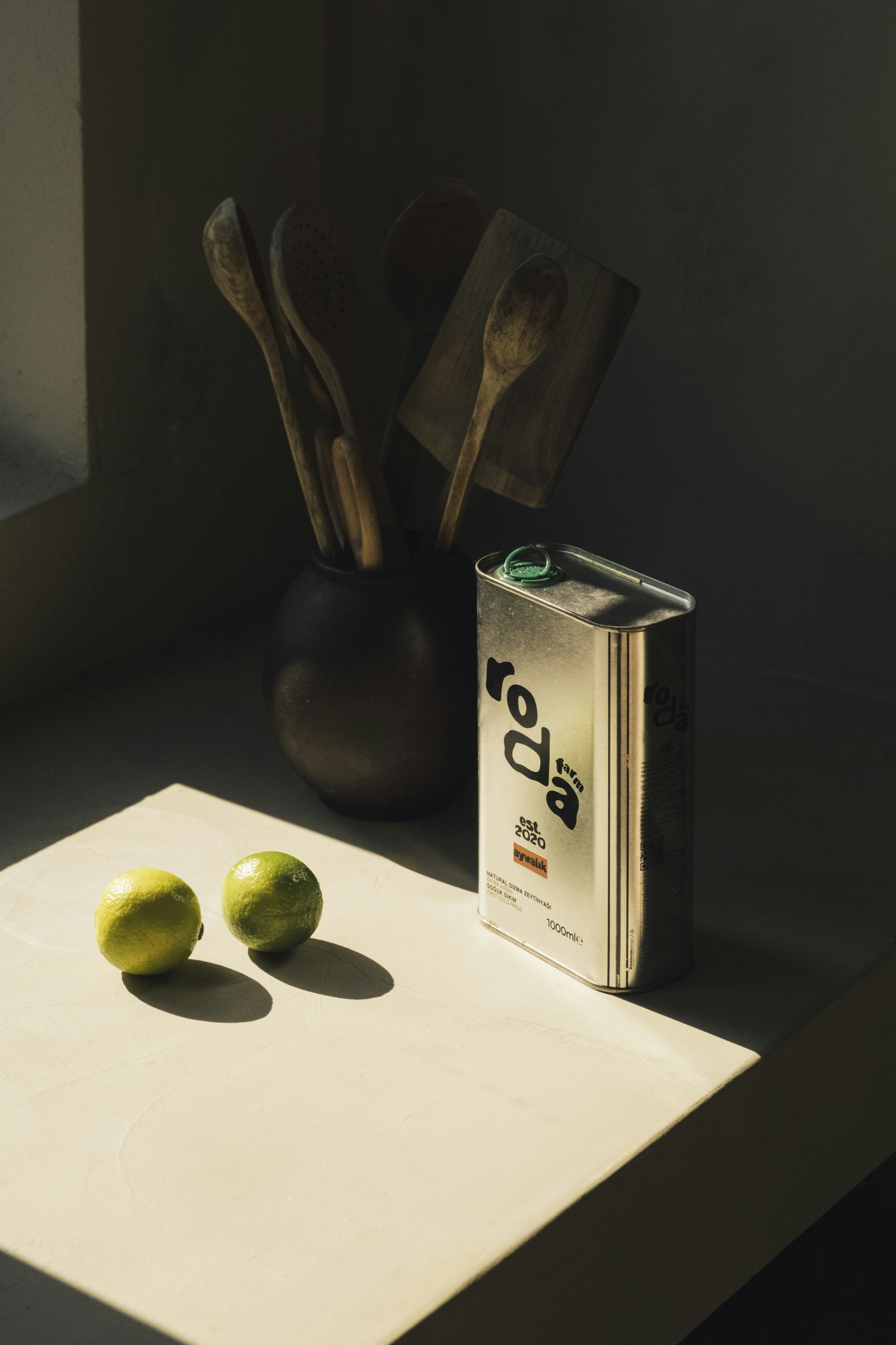 Two limes and a sleek oil canister on a sunlit countertop, accompanied by wooden utensils in the background. The composition highlights the interplay of light and shadow.