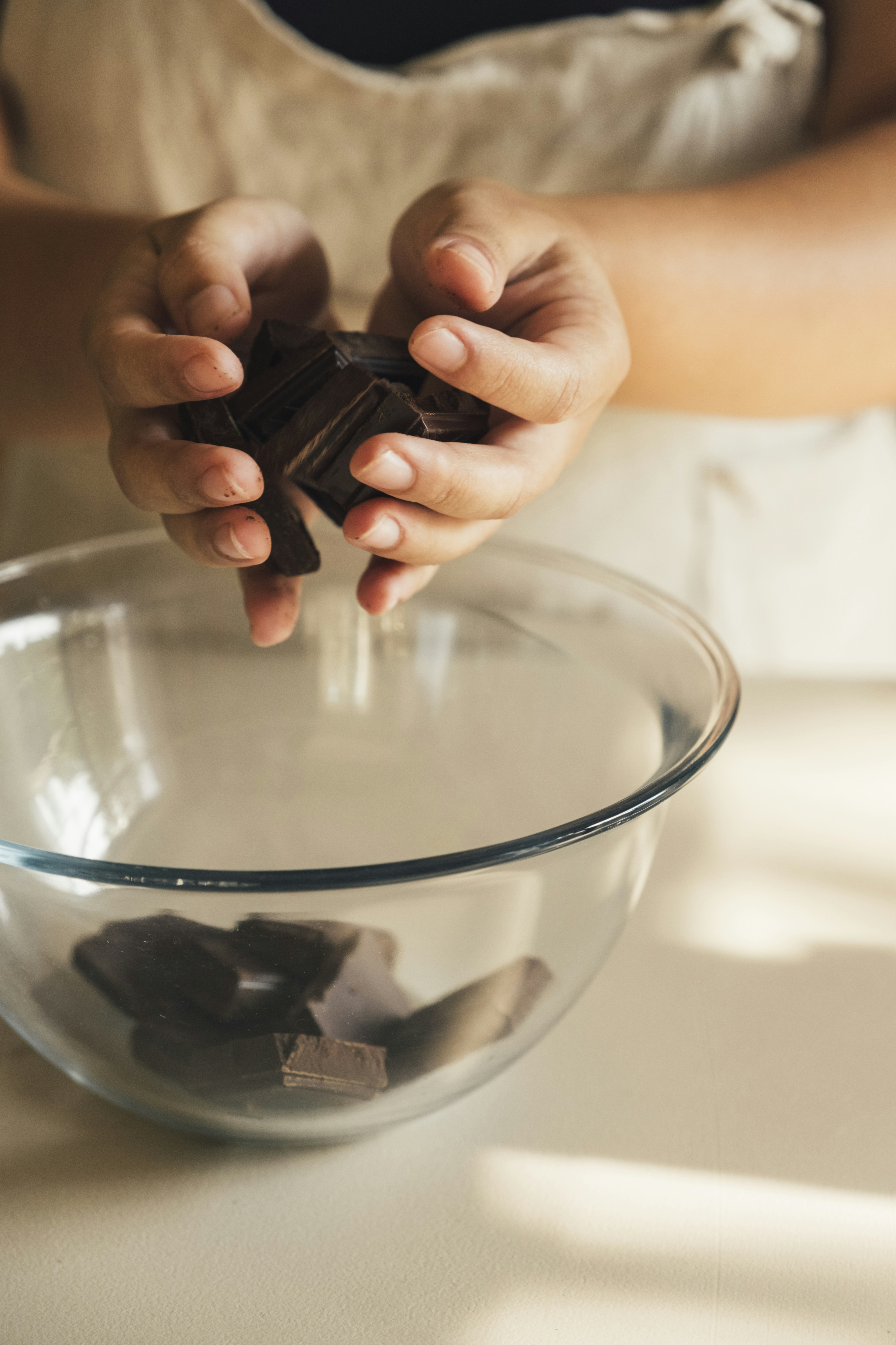 Hands breaking chocolate pieces over a clear glass bowl, ready for baking. Soft light highlights the textures of the chocolate and hands.