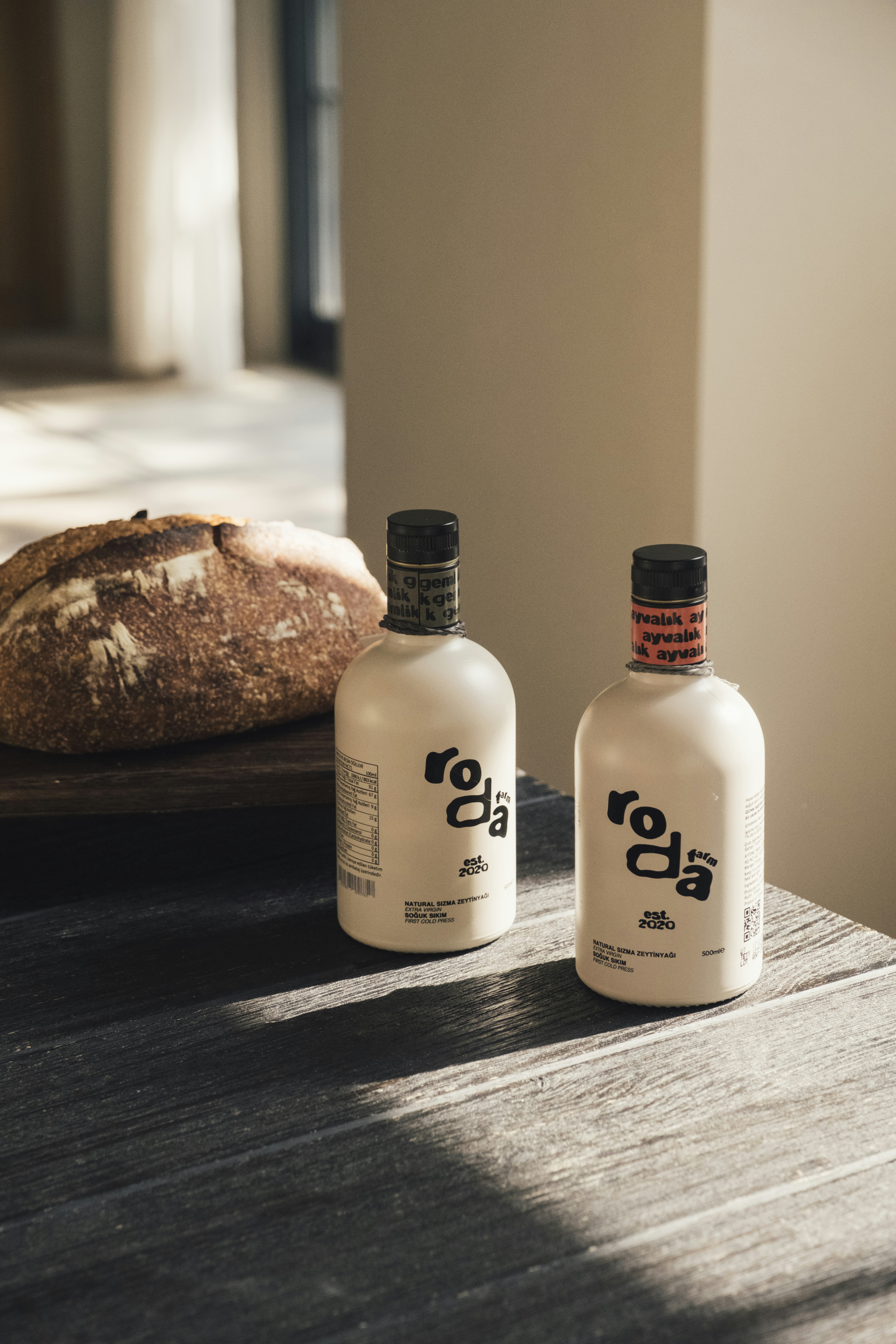 Two elegantly designed bottles of a beverage stand beside a rustic loaf of bread on a textured wooden table, illuminated by soft natural light.