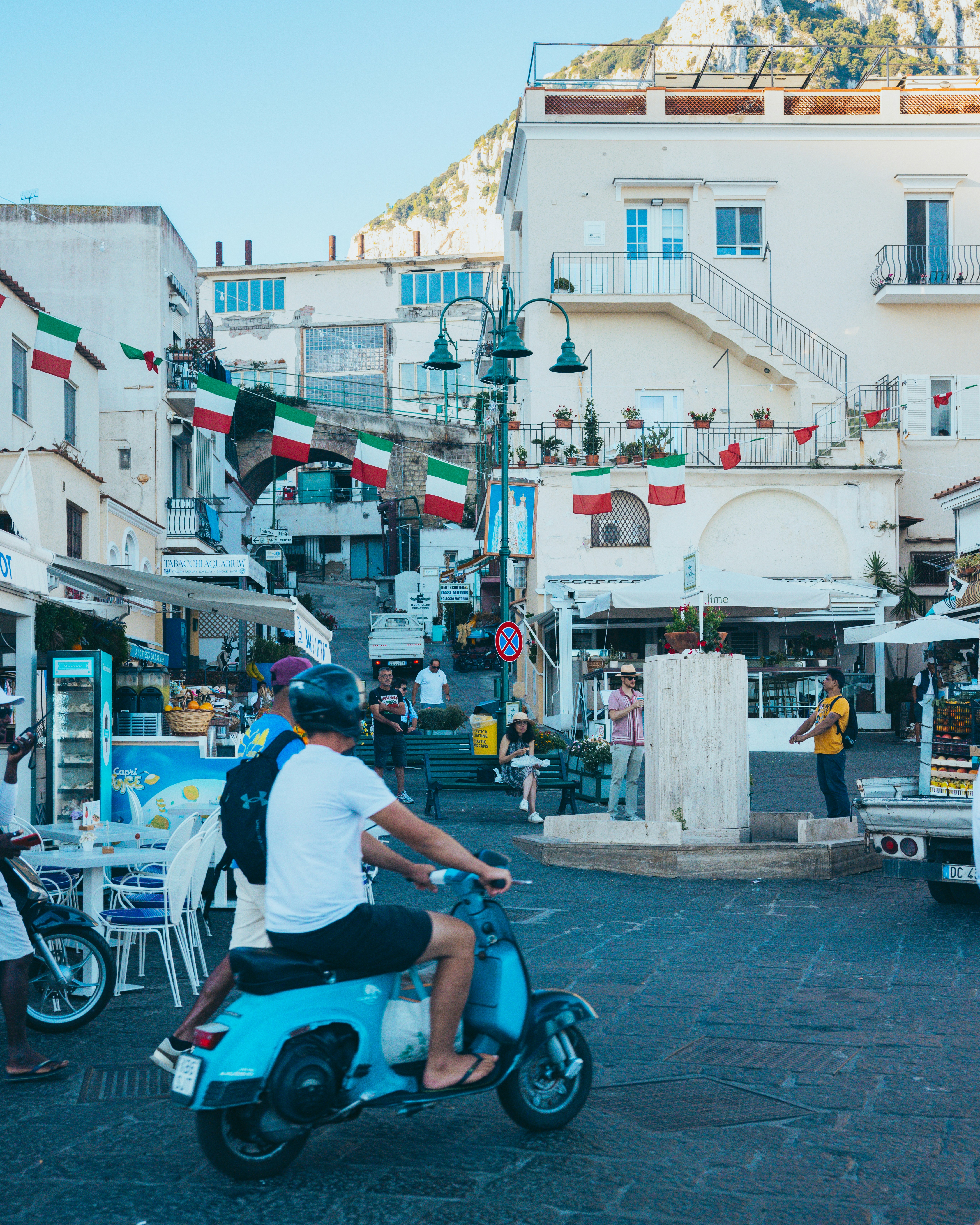 A bustling street scene in a quaint Italian town, featuring a rider on a blue Vespa amidst vibrant flags and lively shops.