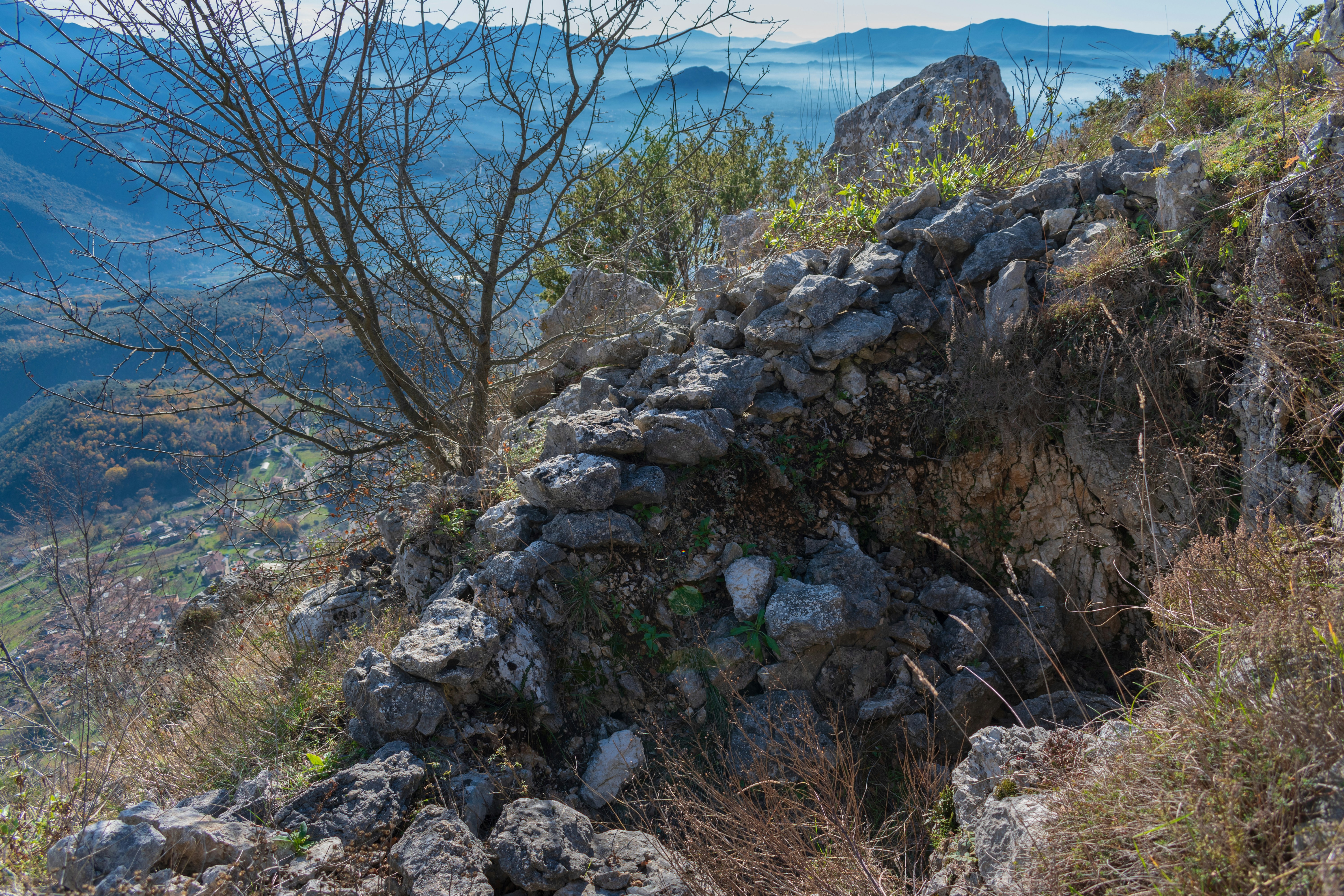 Weathered stone ruins partially covered by grass and vegetation, set against a backdrop of rolling hills and distant mountains.