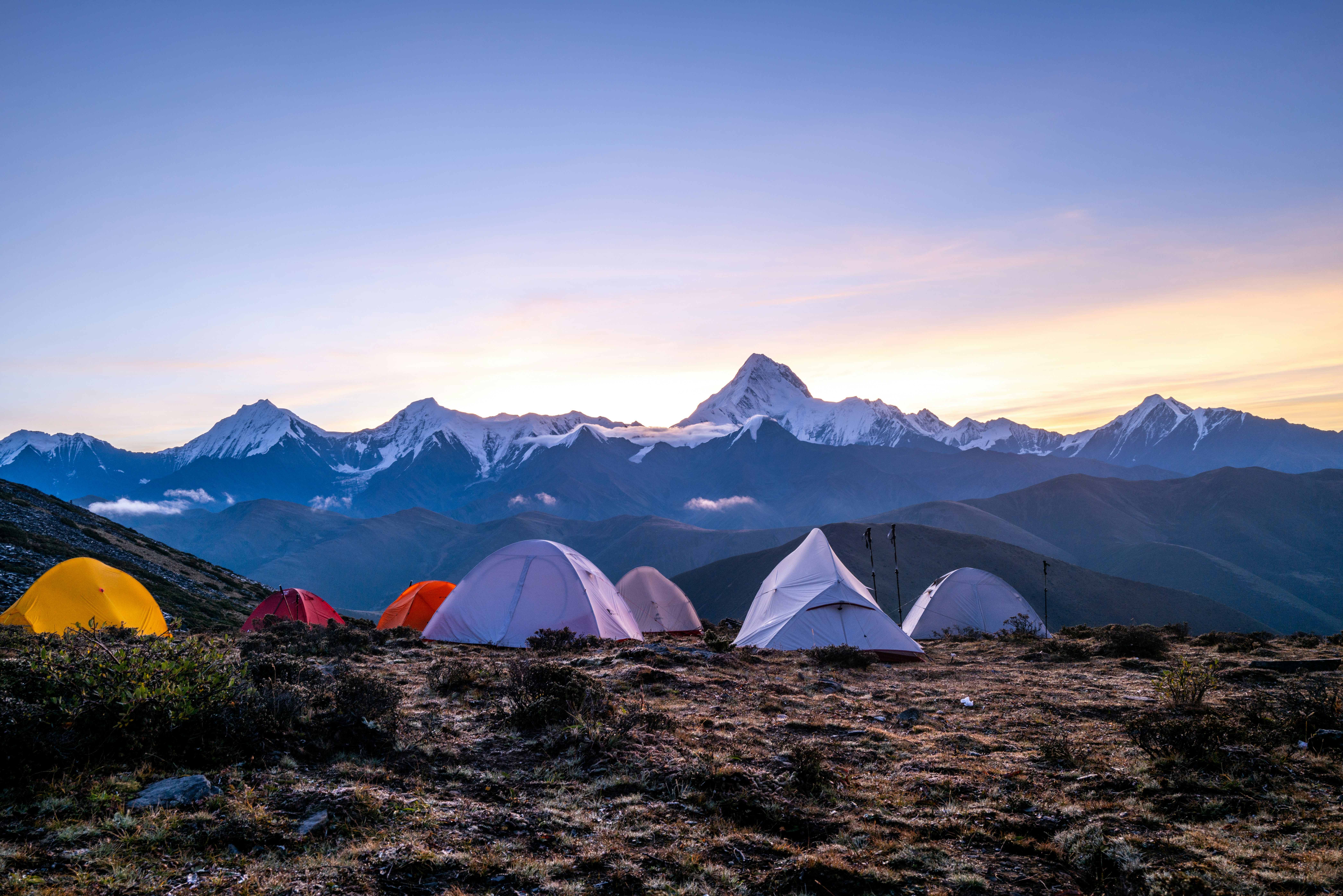 Colorful tents nestled on a mountain ridge as dawn breaks, with majestic snow-capped peaks in the background.