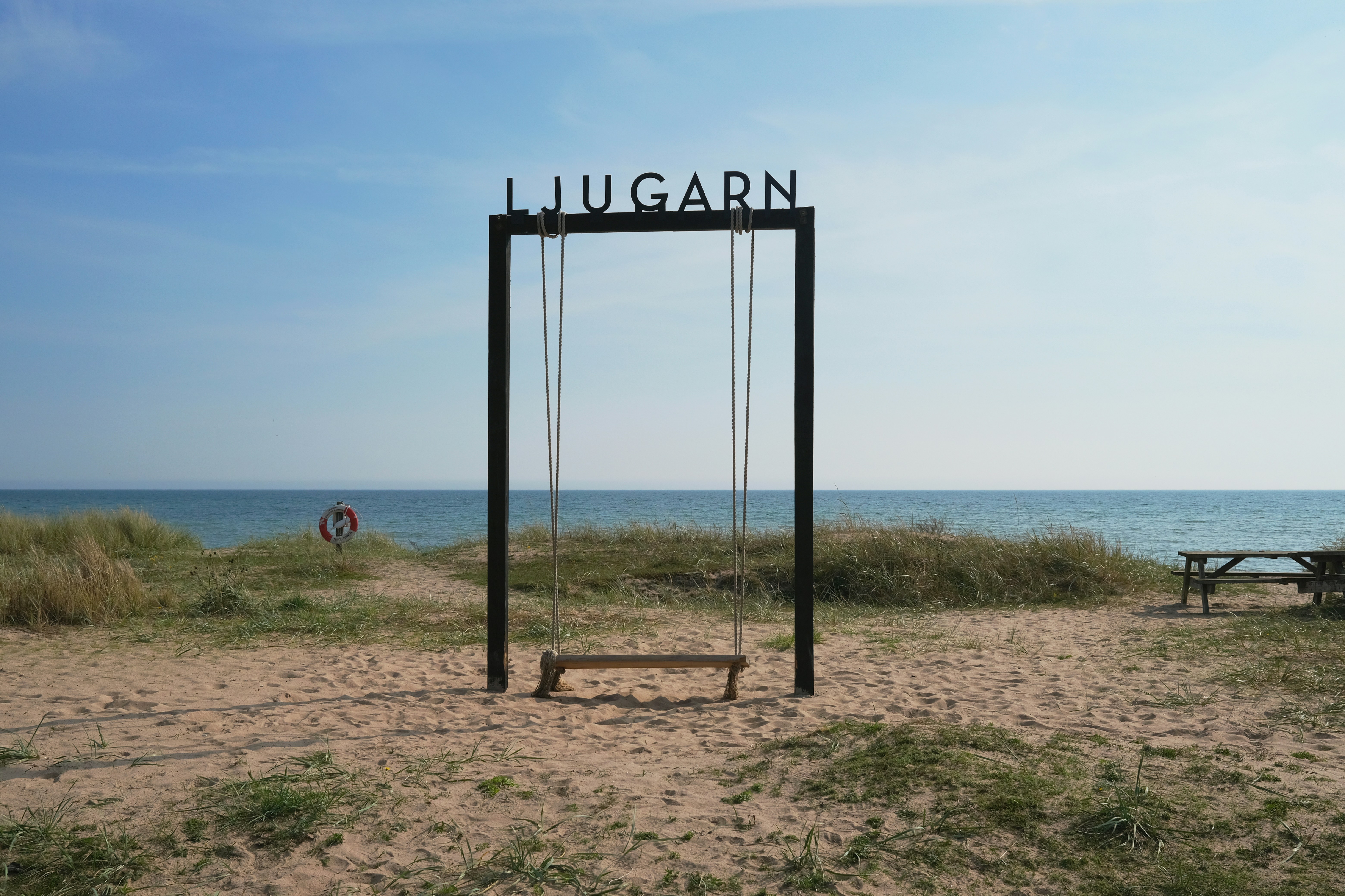 A swing at the beach on sunny day with the Baltic Sea in the background. The name Ljugarn is on the top of the swing. | Swing set on a sandy beach with ocean background