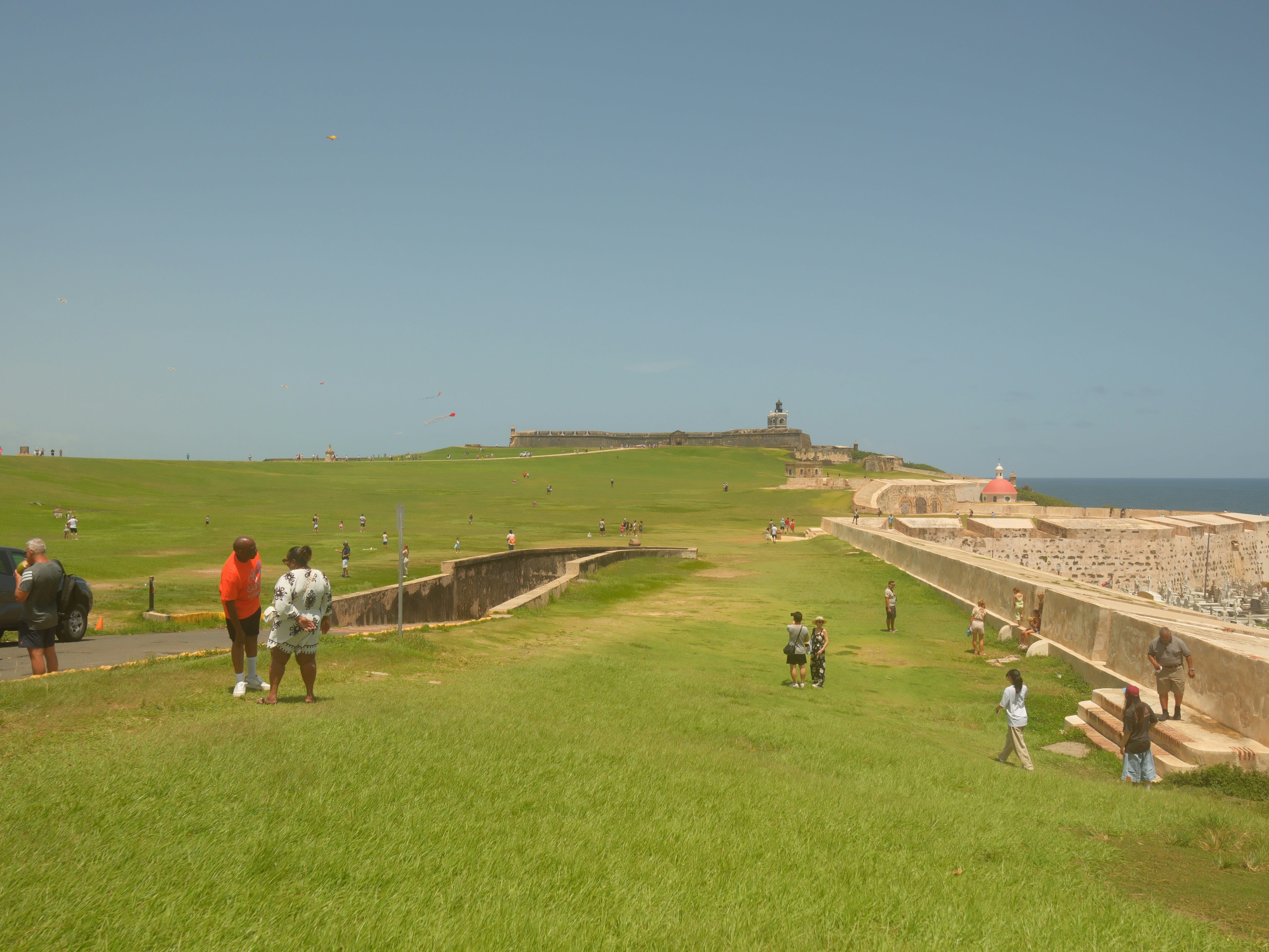 Visitors stroll across lush green grounds leading to a historic fort overlooking the ocean. The scene captures a blend of leisure and heritage.