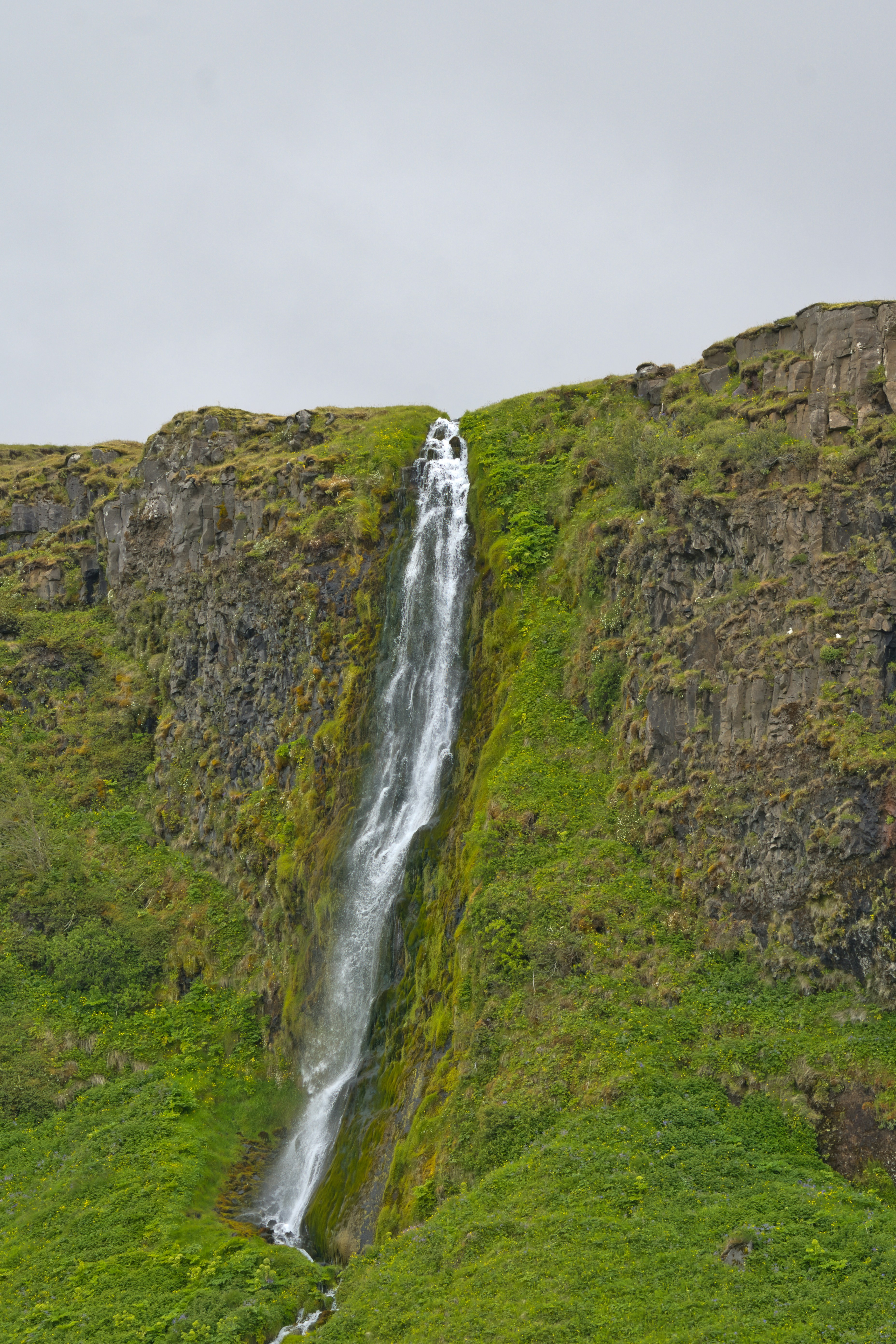 A slender waterfall cascades down a lush green cliff, surrounded by rocky terrain and vibrant vegetation.