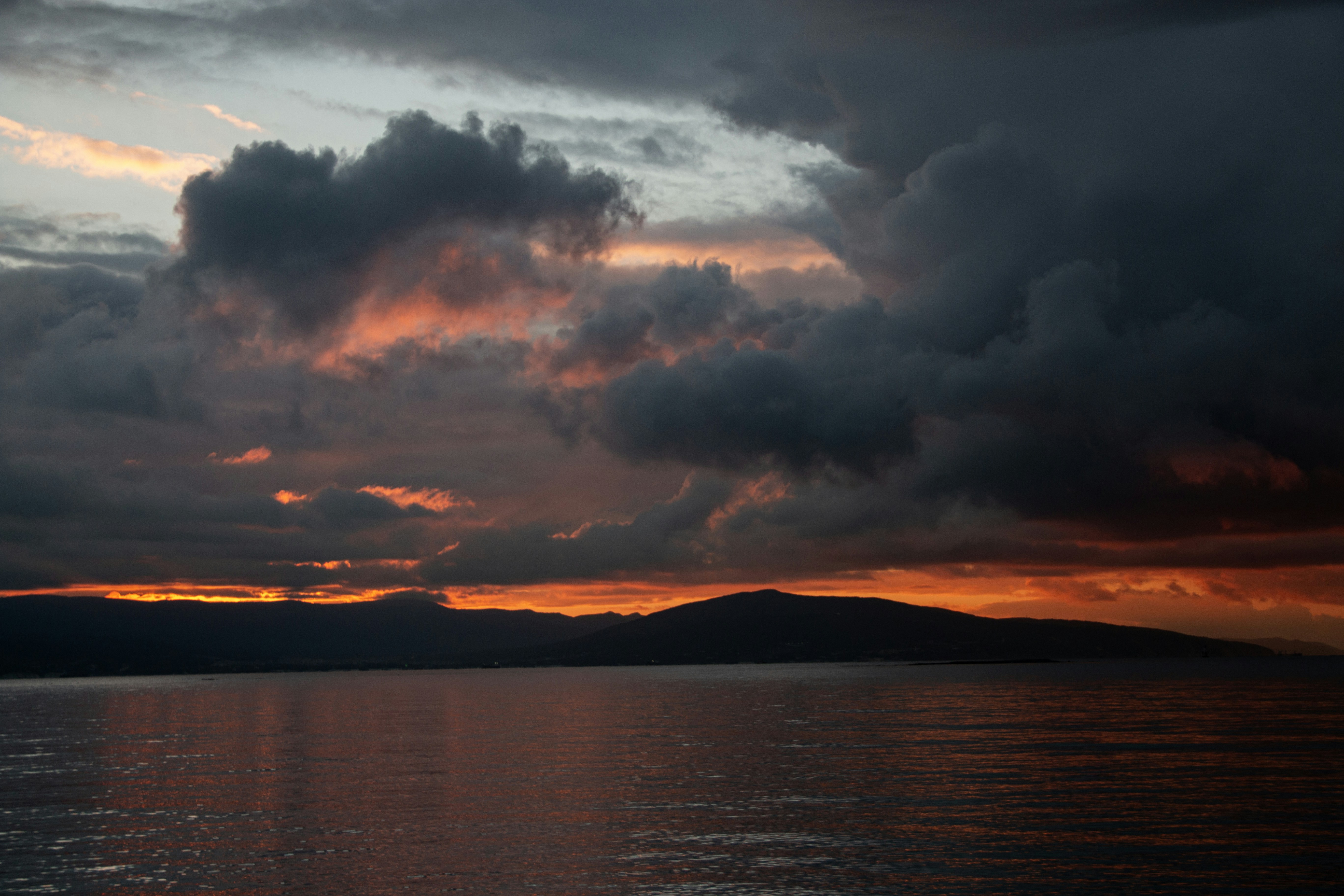 Dramatic clouds gather as the sun sets, casting warm hues over a tranquil sea and distant hills. The interplay of light and shadow creates a mesmerizing atmosphere.