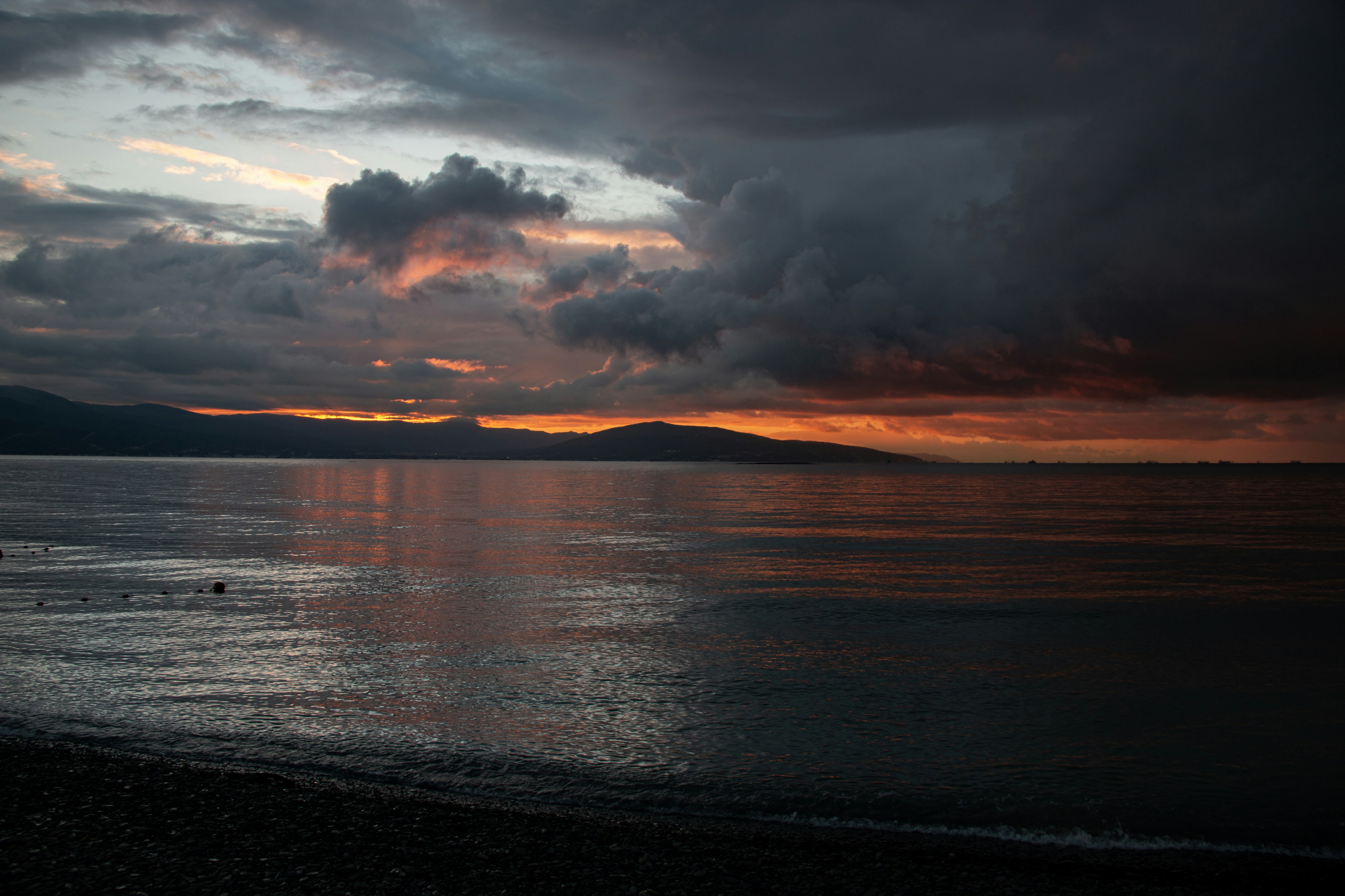 Dark clouds loom over a serene bay, reflecting vibrant hues of orange and purple on the water's surface. The distant hills frame the scene, enhancing the atmospheric mood.