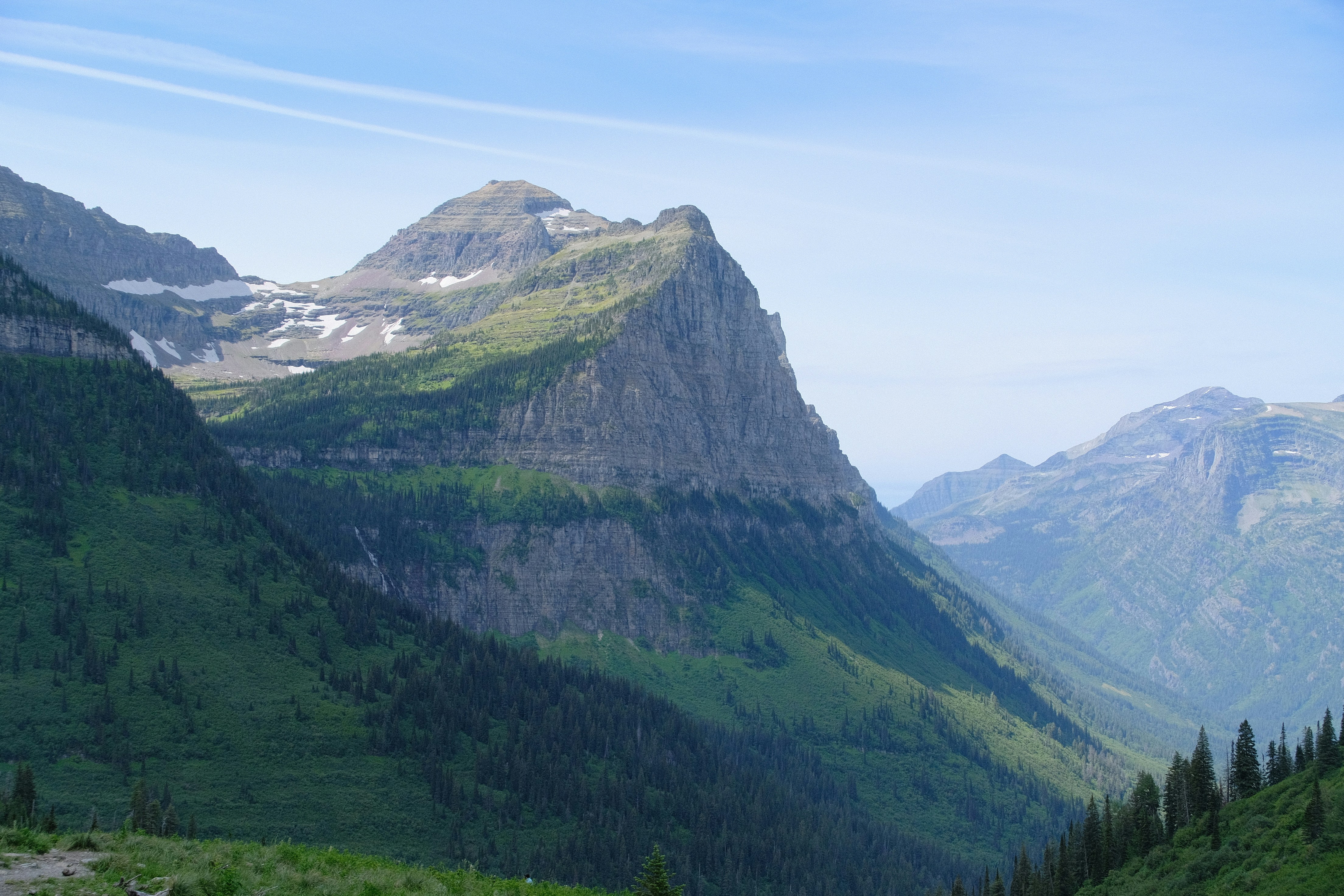 Lush green valley with towering mountains and patches of snow under a clear blue sky.