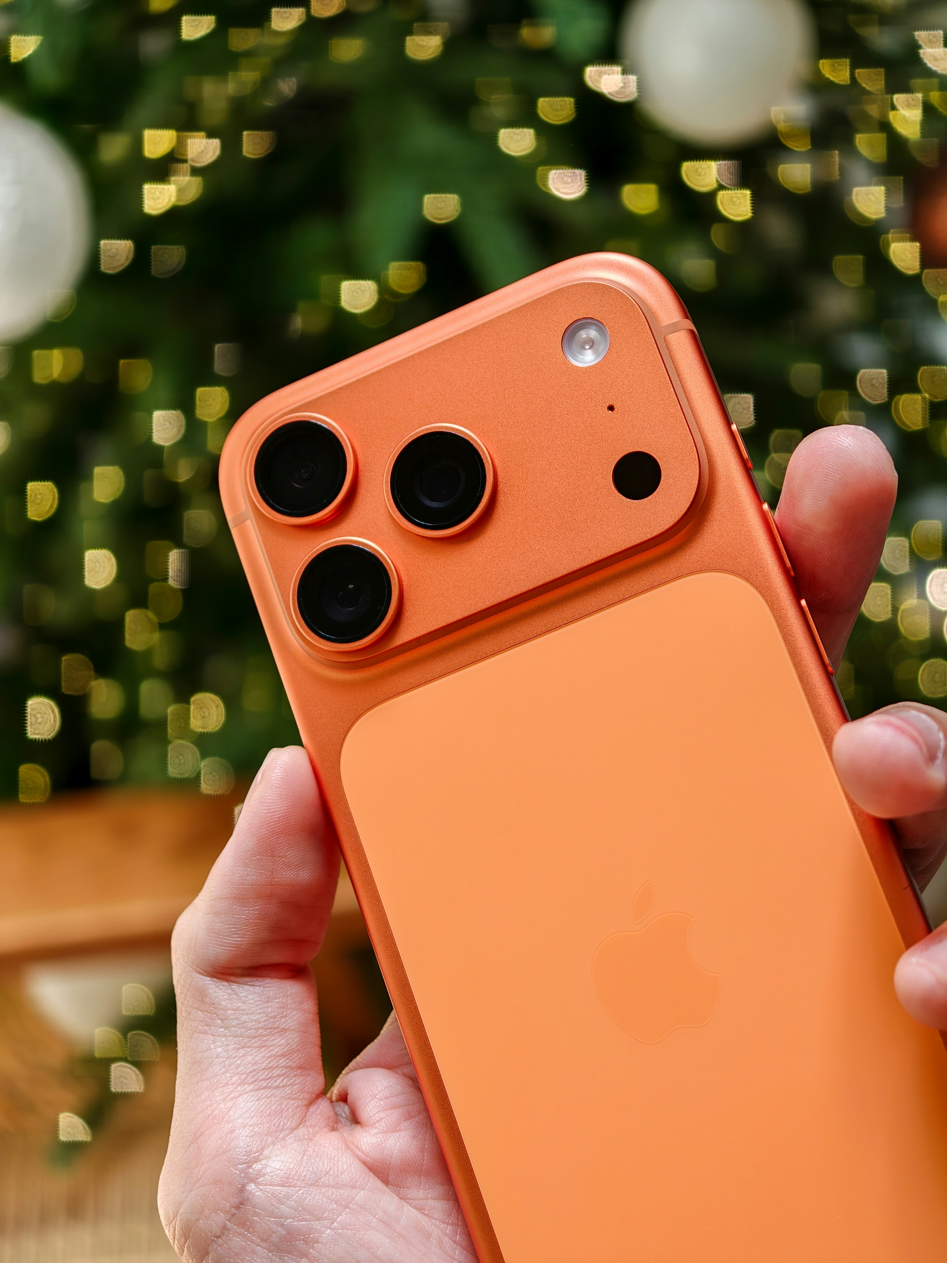 Apple iPhone 17 Pro Cosmic Orange | Hand holding an orange smartphone with christmas tree bokeh