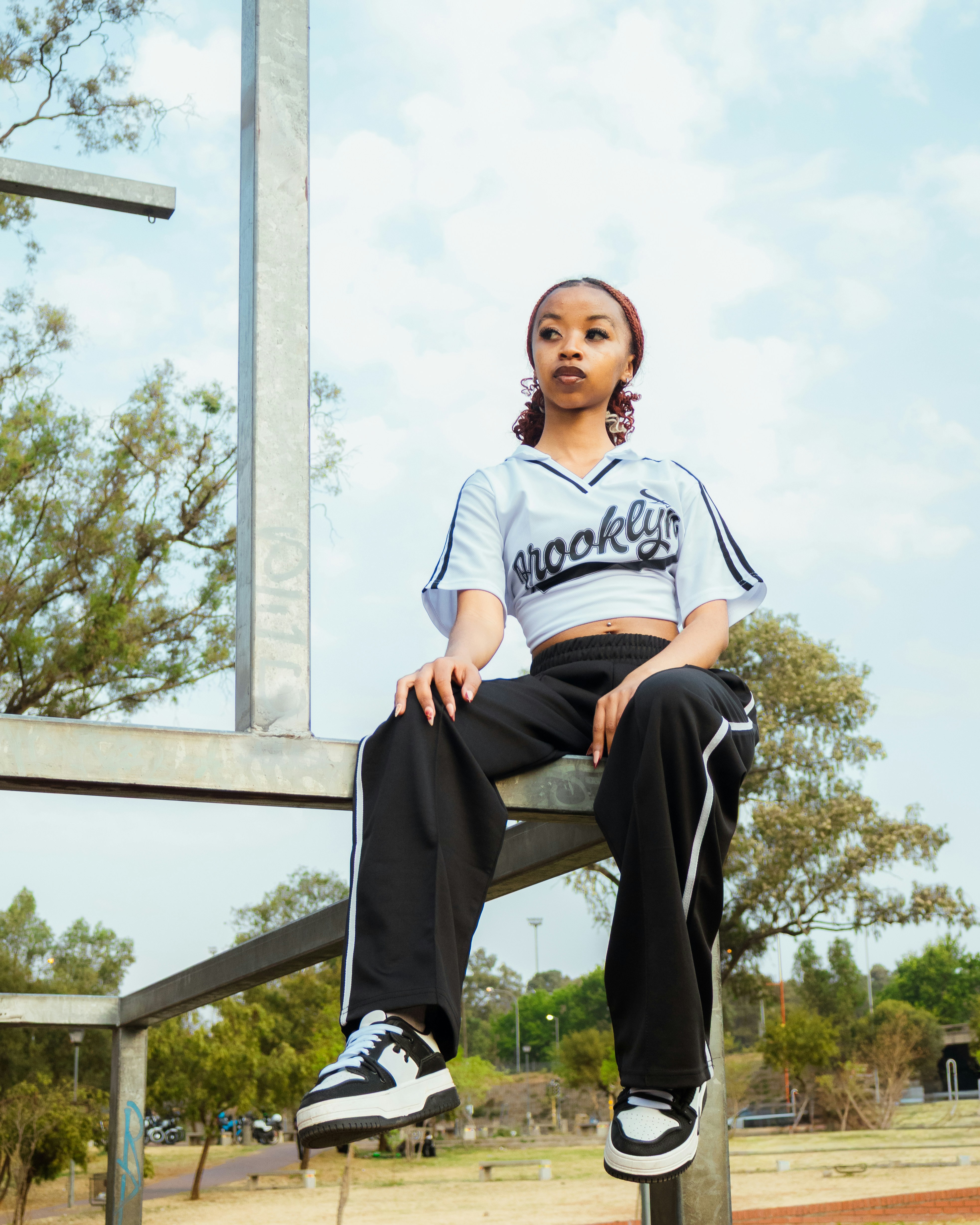 A young woman casually poses on a metal structure in a park, showcasing a sporty outfit with a Brooklyn-themed top and stylish sneakers.