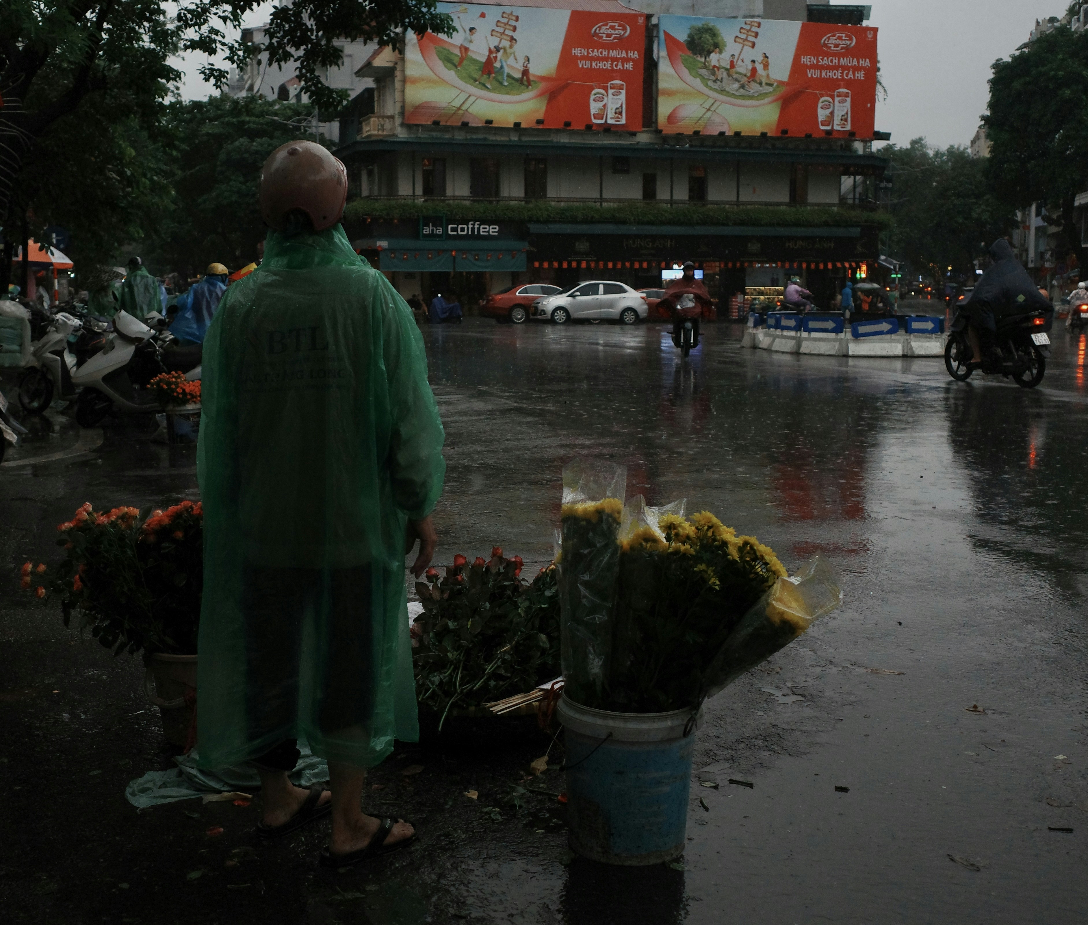 Person in green raincoat stands by flower stall in rain.