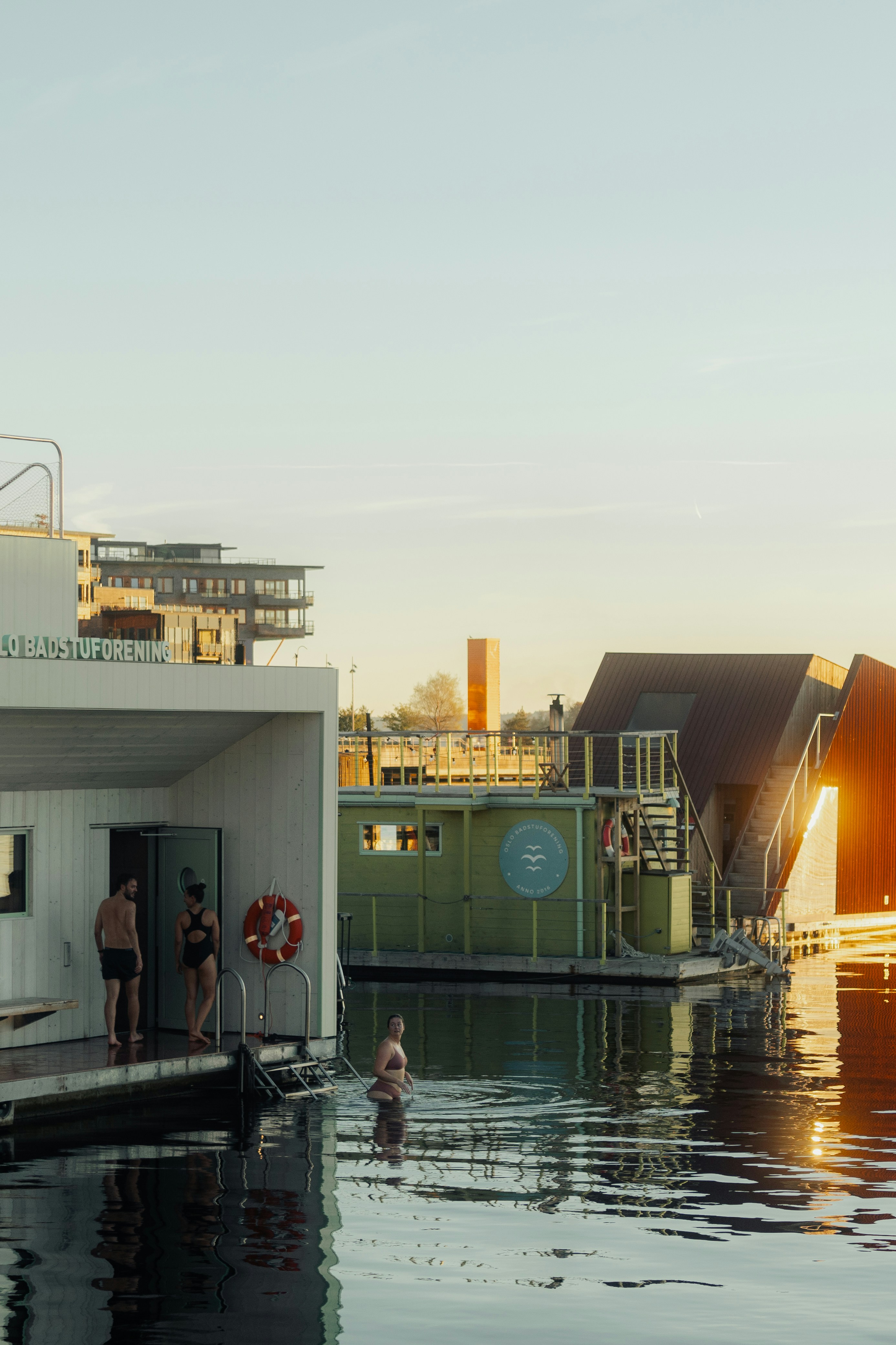 Bathers enjoying the serene waterside atmosphere near modern architecture and a floating sauna. The warm light enhances the tranquil setting.