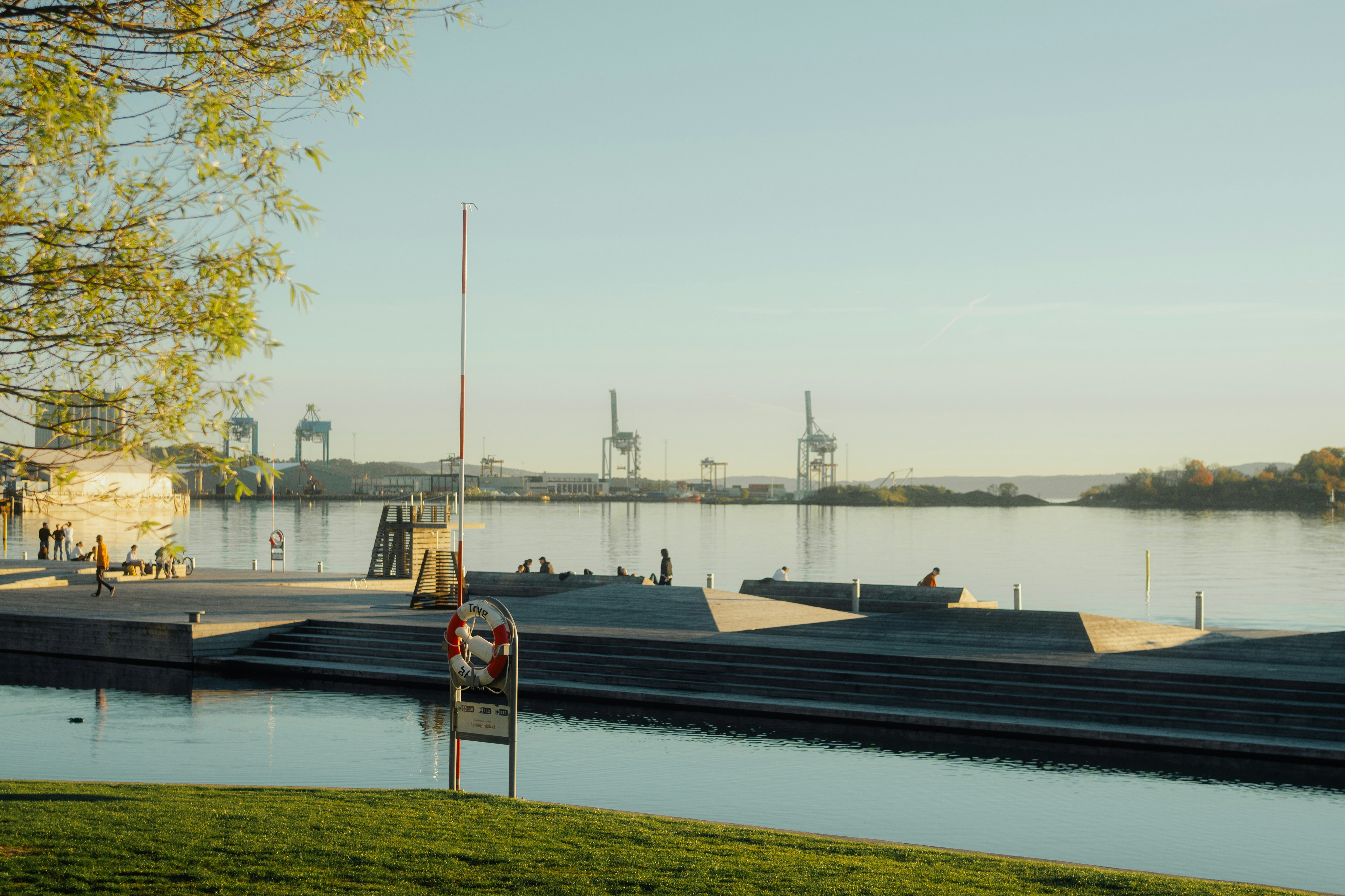 A peaceful waterfront scene featuring a modern harbor with cranes in the background, reflecting calm waters and a clear sky. People enjoy leisure time along the promenade.