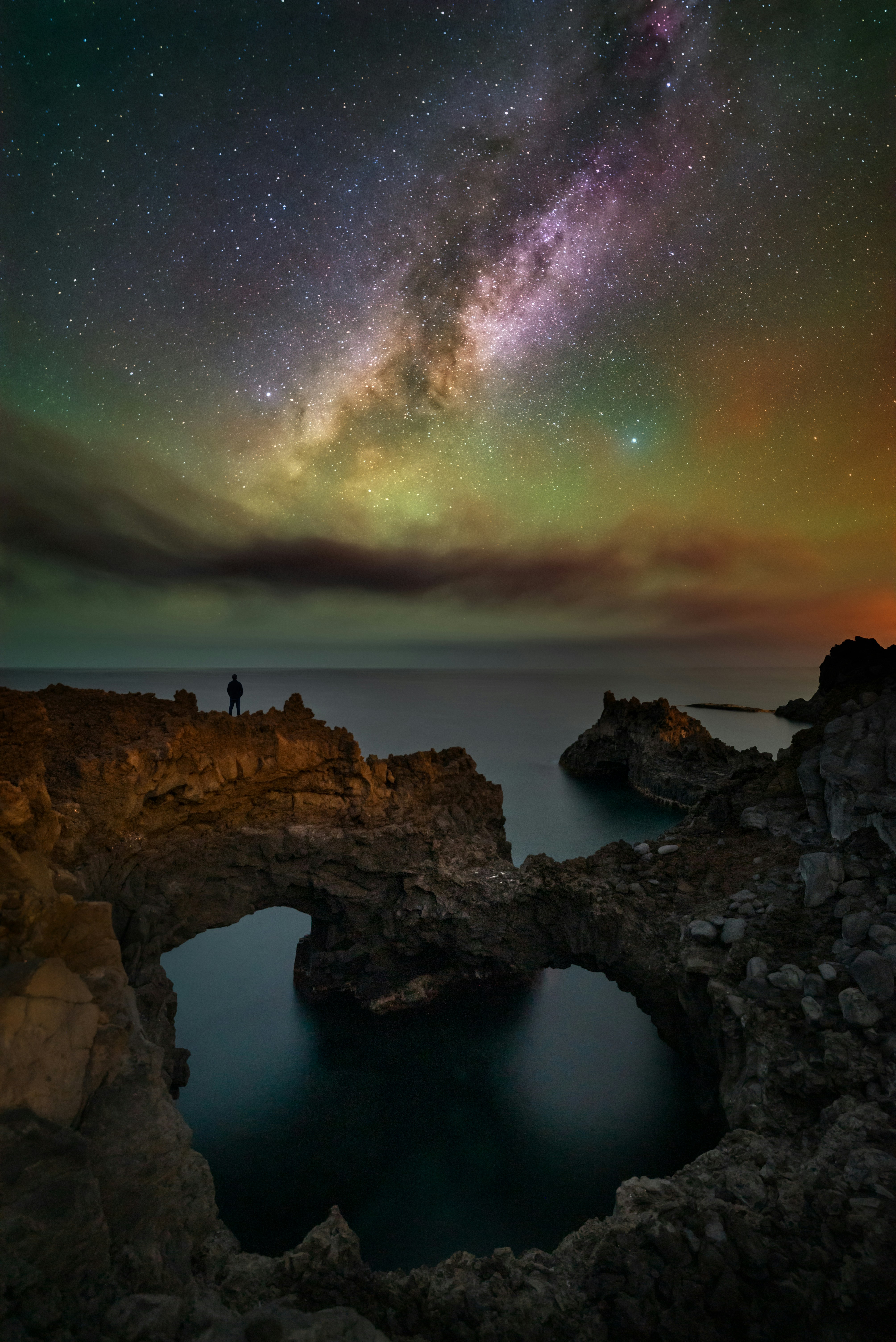 Man stands on rocky shore under starry night sky.
