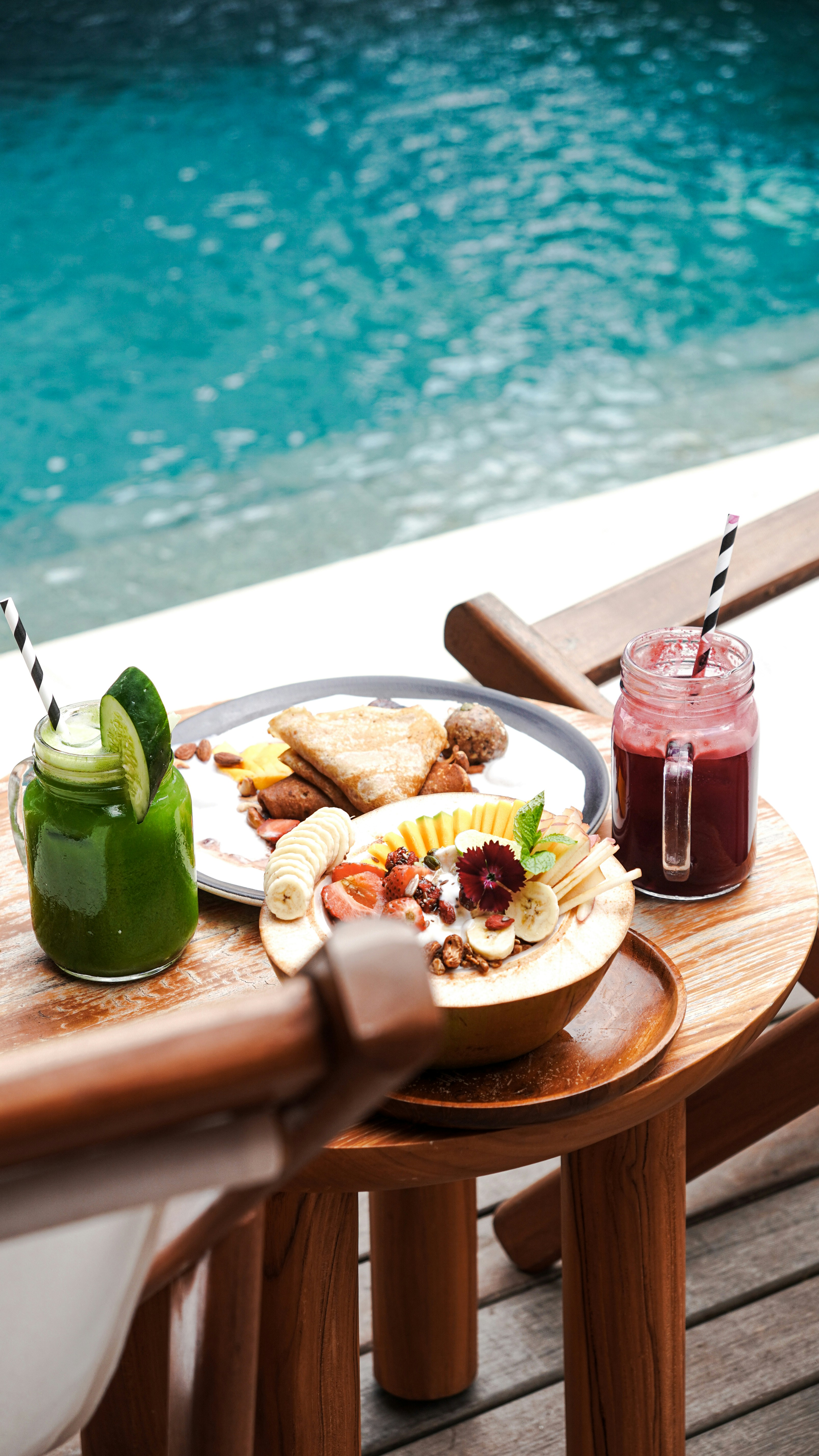 A vibrant spread of tropical fruits and beverages on a wooden table beside a serene blue pool. The scene invites relaxation and enjoyment.