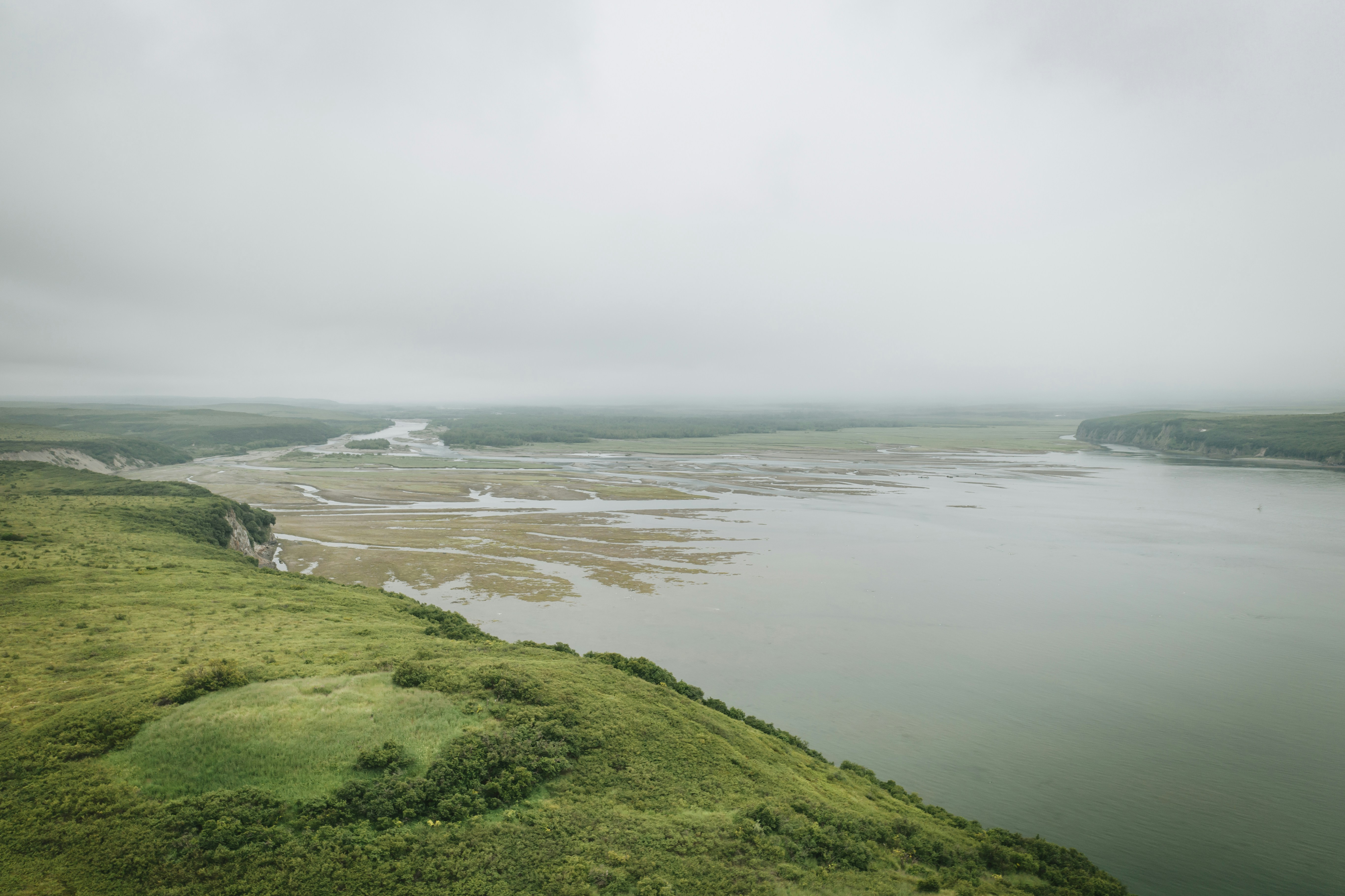 Expansive view of a tranquil estuary surrounded by lush greenery under a cloudy sky. The gentle flow of water meanders through the landscape.