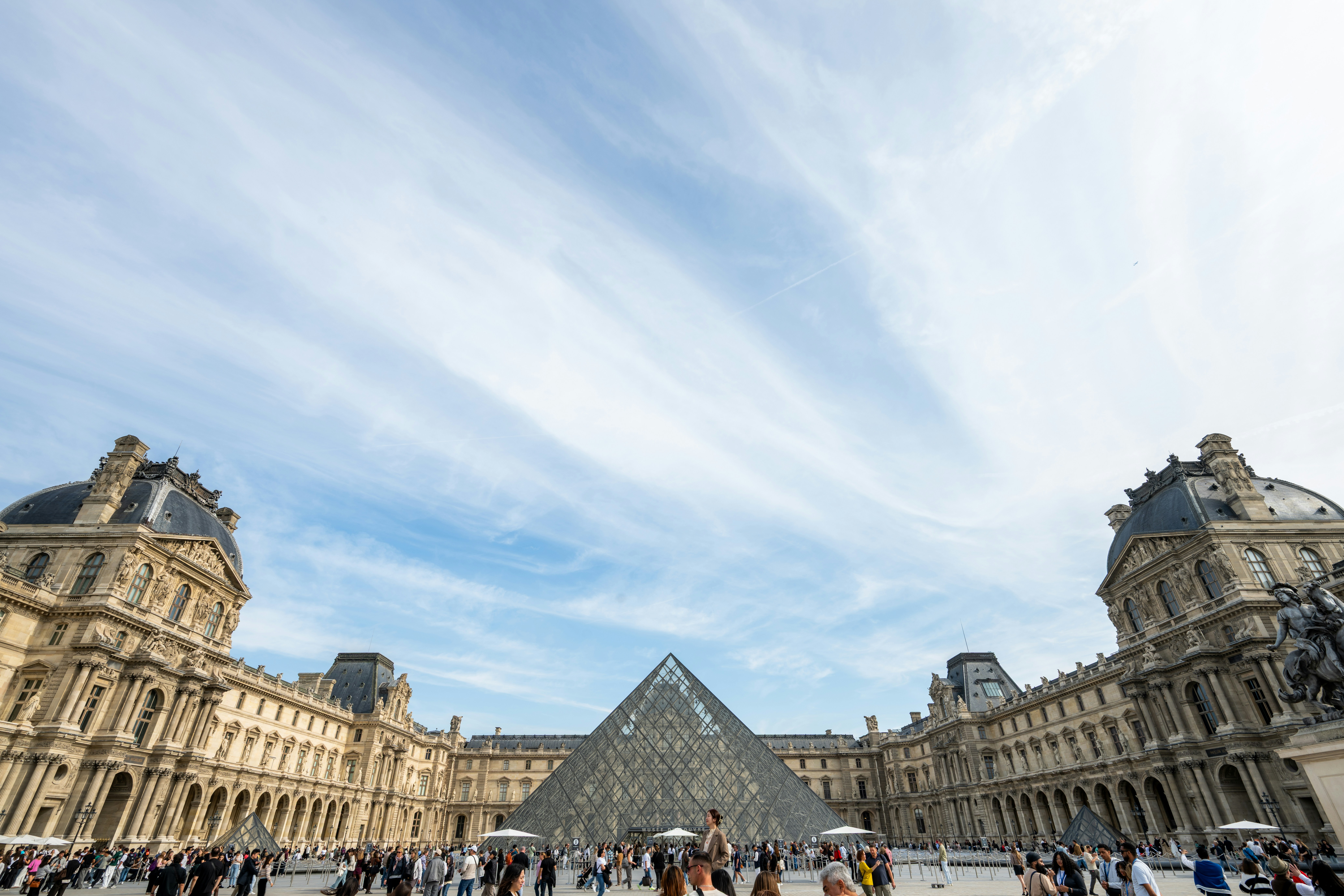 Iconic glass pyramid of the Louvre surrounded by historic architecture and bustling crowds. A blend of modern design and classic elegance.