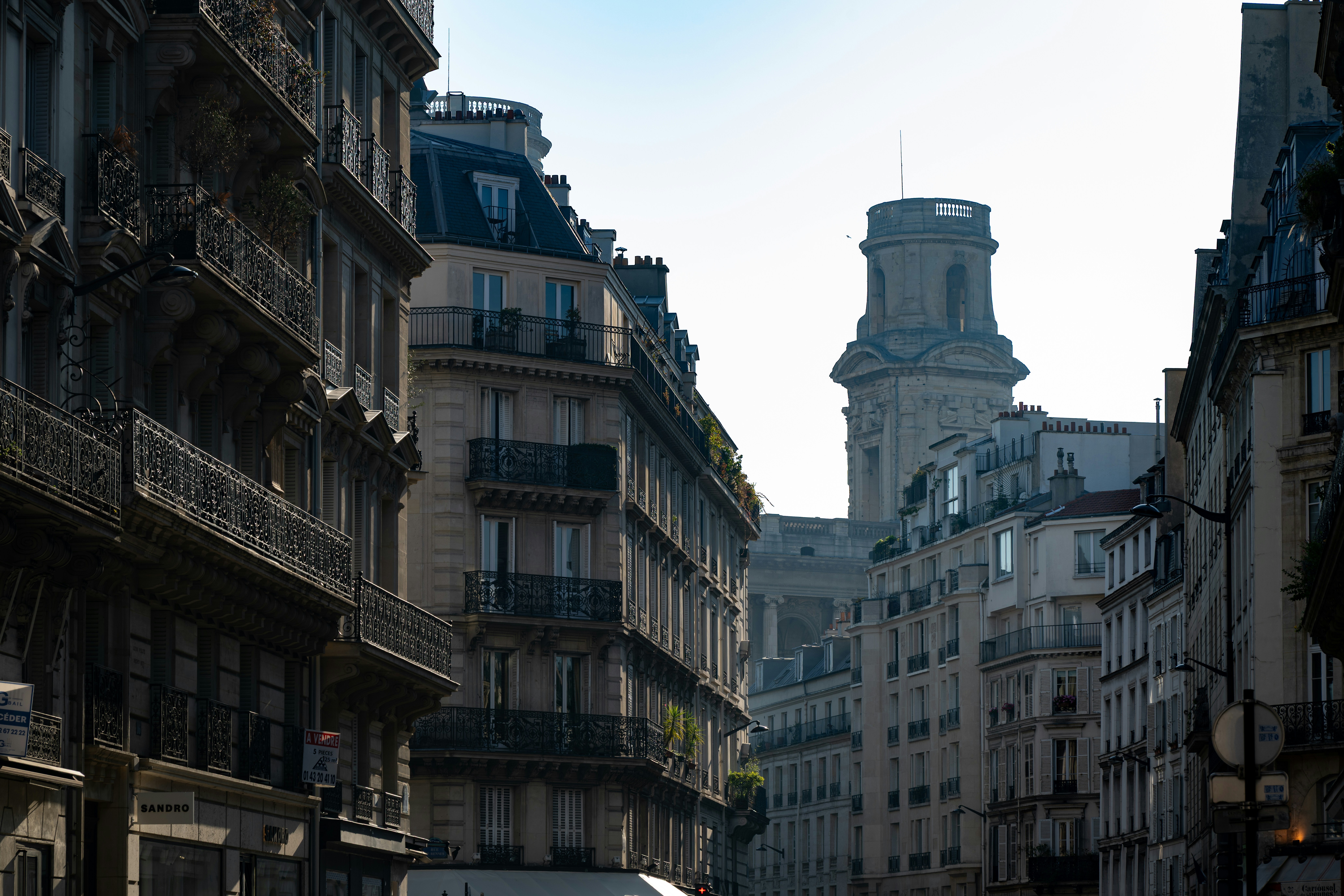 Historic Parisian buildings line a narrow street, leading to a distant tower under a clear sky.