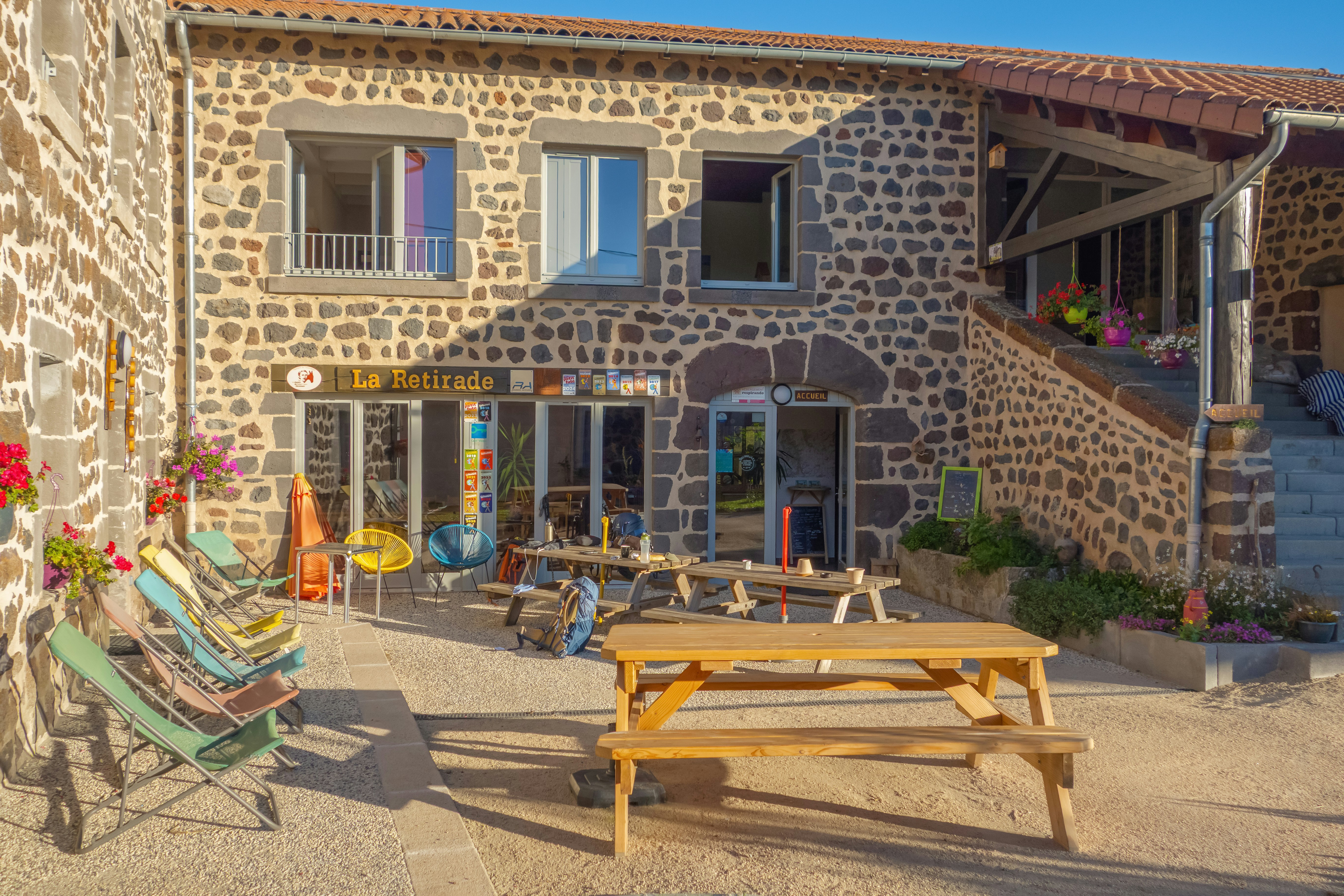 A cozy courtyard featuring a rustic stone building, colorful seating, and vibrant flowers, inviting relaxation and social gatherings.