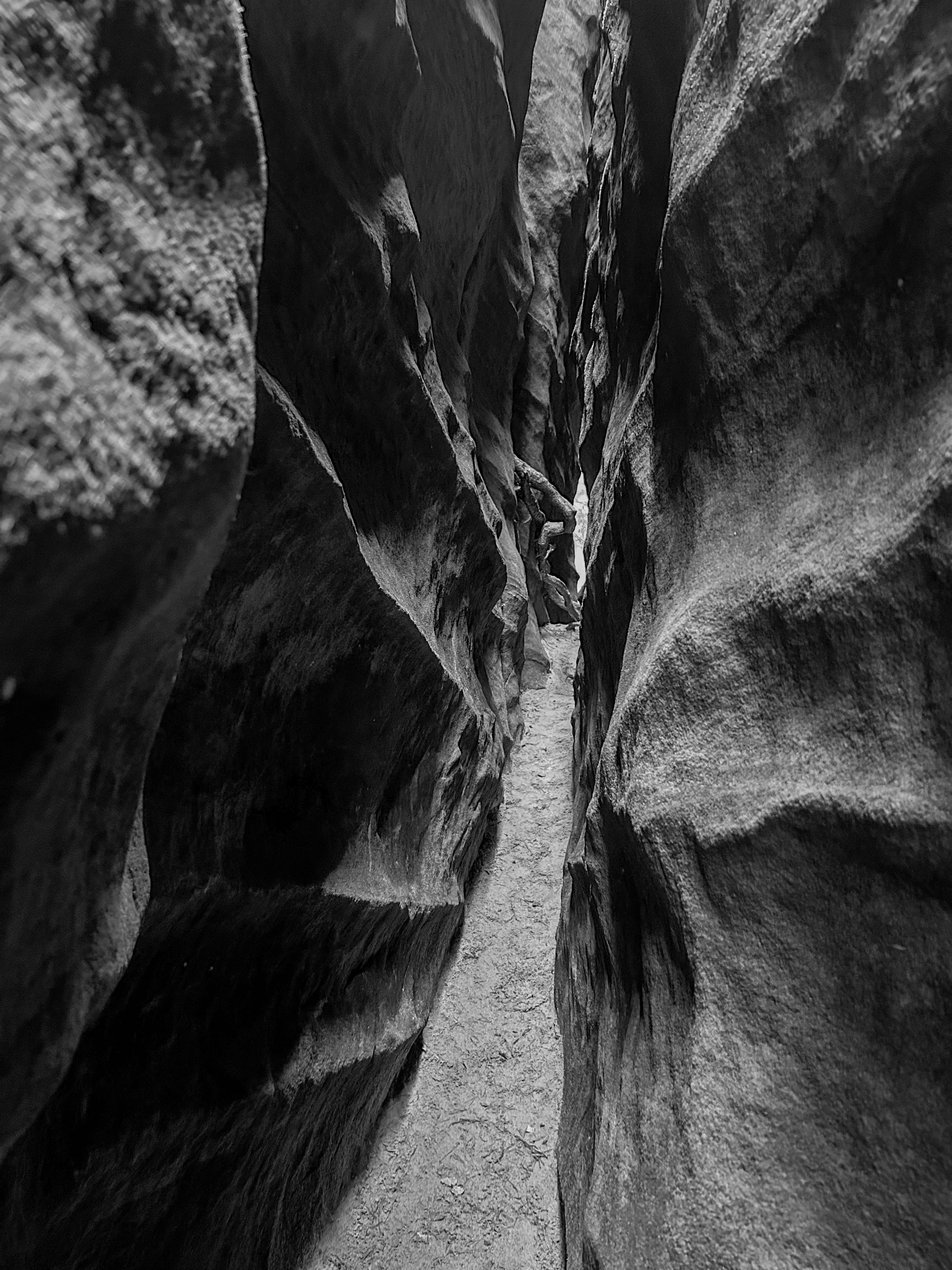 Monochrome photograph of a narrow sandstone canyon passage with textured rock walls and a sandy path. The dramatic light and shadow emphasize the natural patterns and depth of the stone formations, creating an abstract and mysterious landscape scene. | Narrow canyon path with towering rock walls