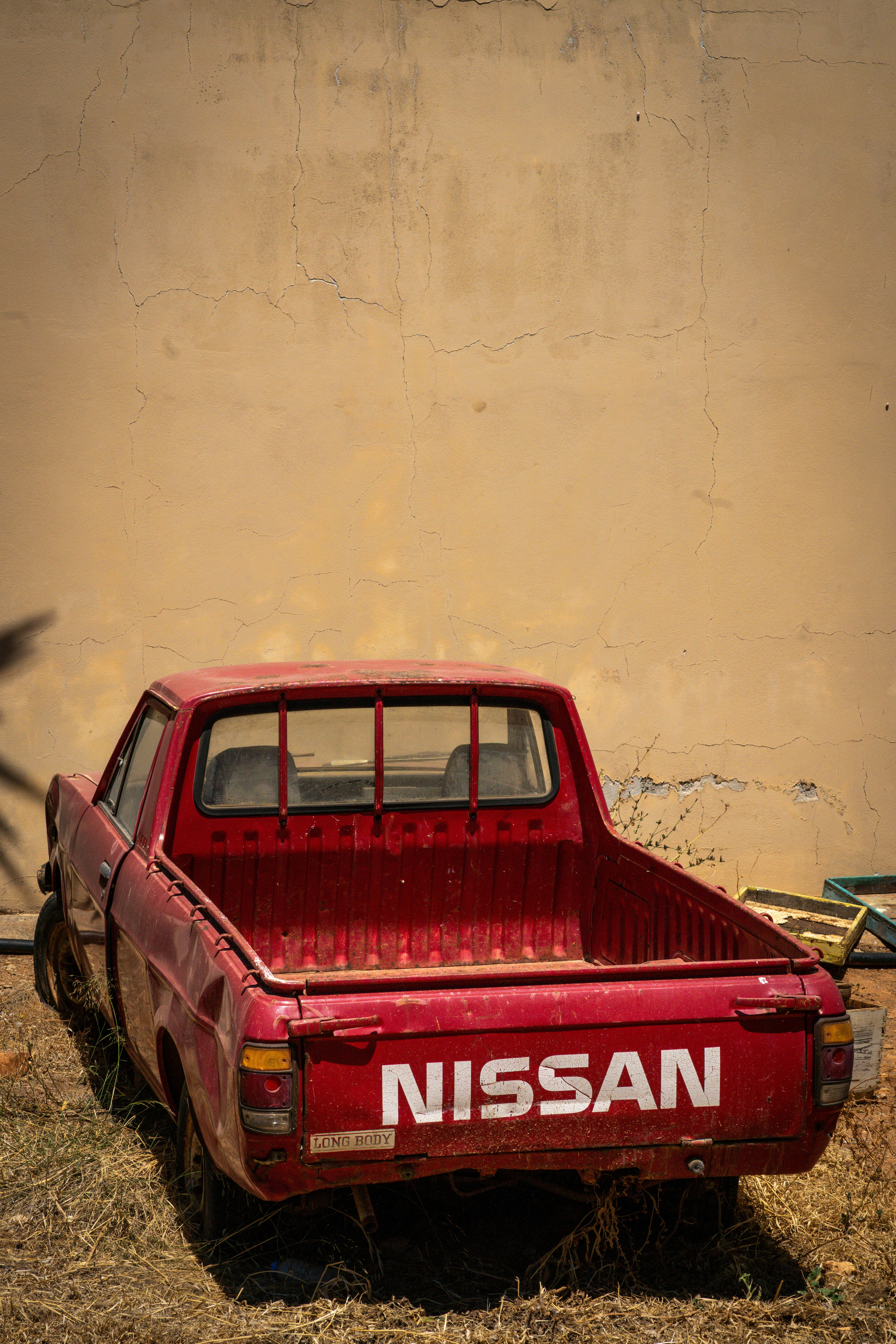 An aged red Nissan pickup truck sits abandoned in a dry, grassy field, its weathered body contrasting against a cracked, sepia-toned wall in the background. The vintage vehicle, with its "Long Body" trim, tells a story of past utility and forgotten journeys. | Old red nissan pickup truck against wall