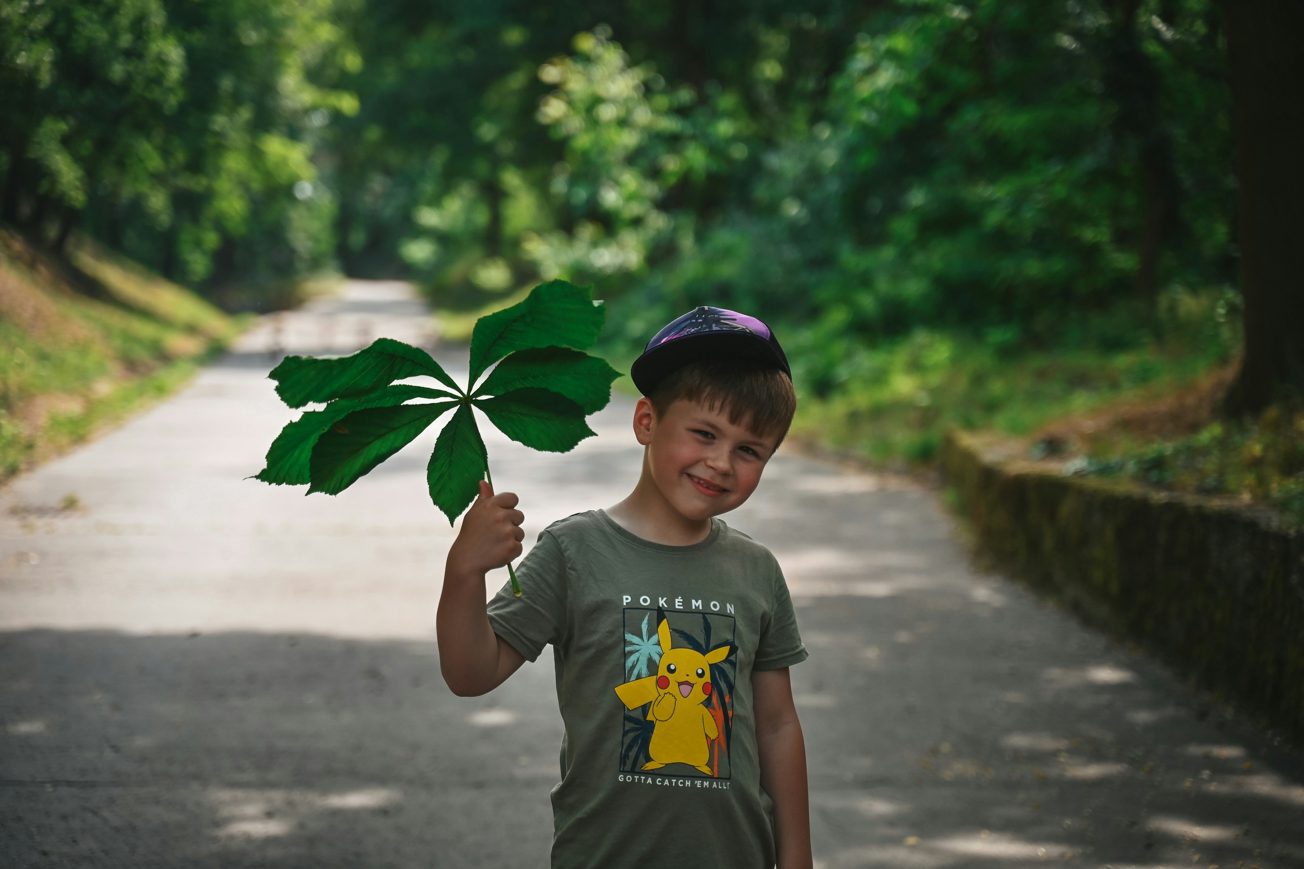 A smiling boy holds a large green leaf outdoors.