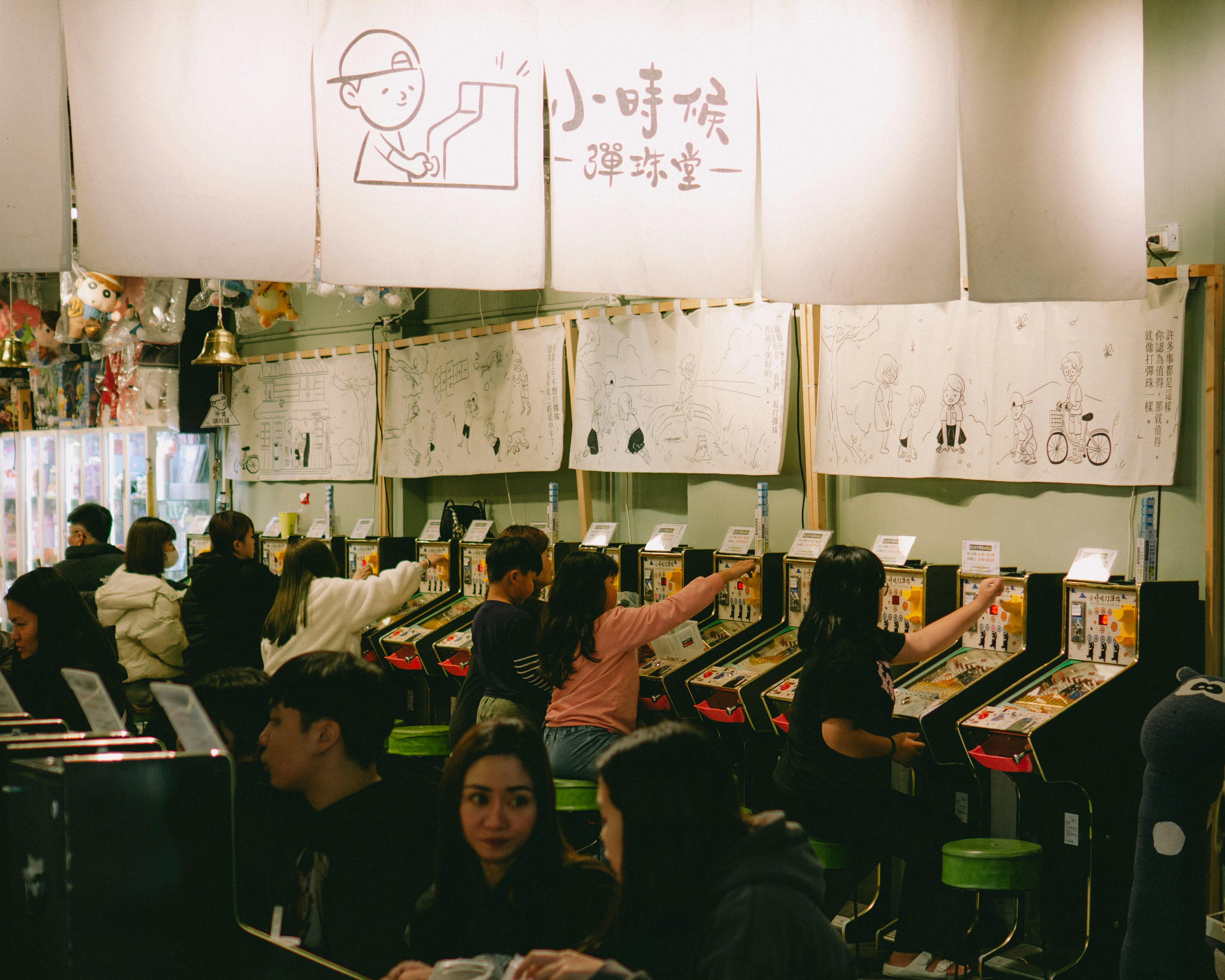 A lively, nostalgic atmosphere fills the 'Childhood Pinball Hall', a retro arcade in Taichung, Taiwan. Rows of patrons are engrossed in playing classic, old-school pinball machines, their focused energy creating a social buzz. Above them, charming, hand-drawn style banners add to the vintage, low-tech vibe of the space. The scene captures a beloved local pastime that transcends generations, offering a simple, hands-on form of entertainment that evokes fond memories of simpler times. | People playing arcade games in a dimly lit room.