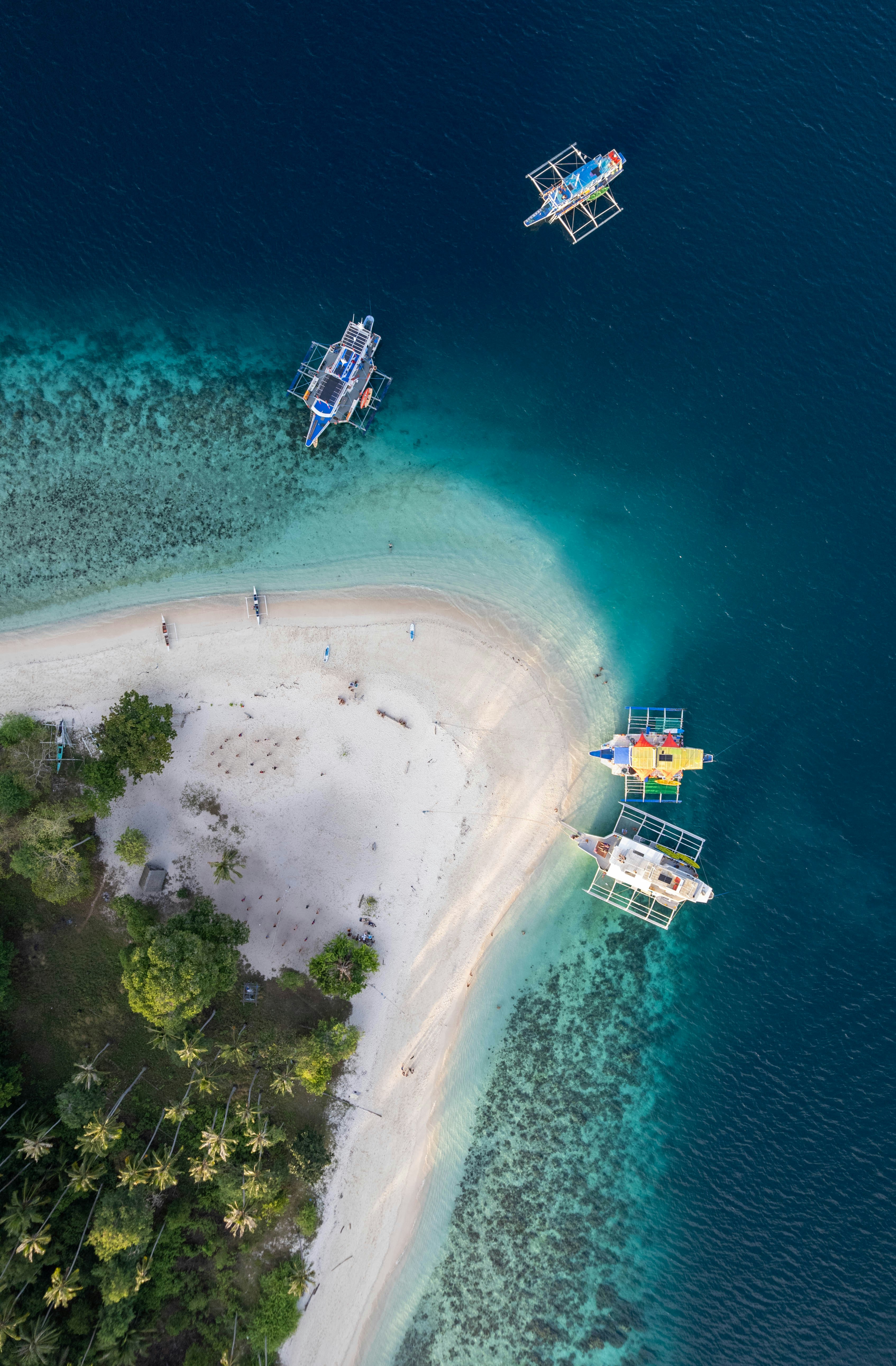 Aerial view of boats anchored near a sandy beach surrounded by turquoise waters and lush greenery.