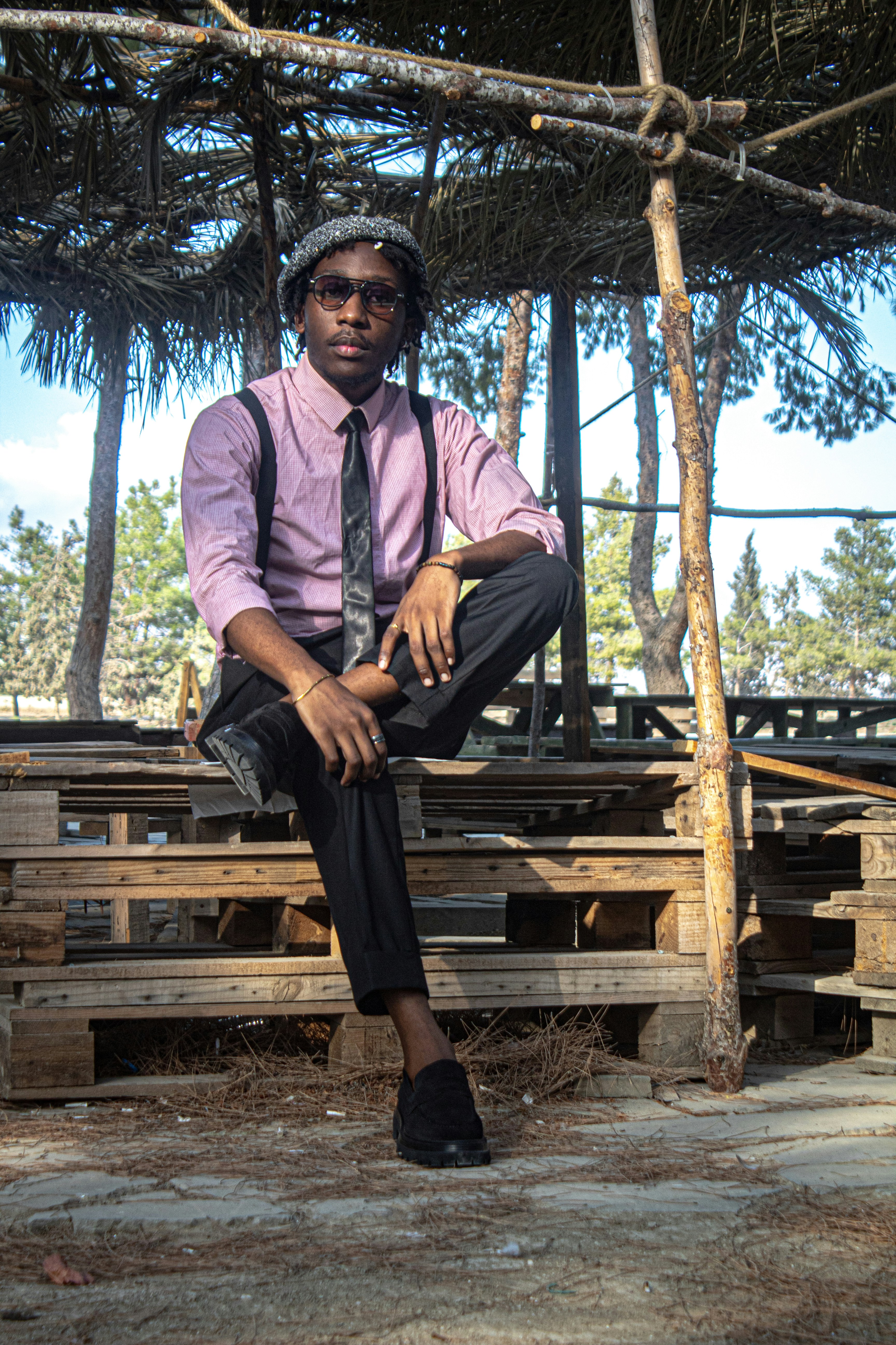 A stylish individual seated on wooden pallets, dressed in a pink shirt and black trousers, surrounded by natural elements and rustic structures.