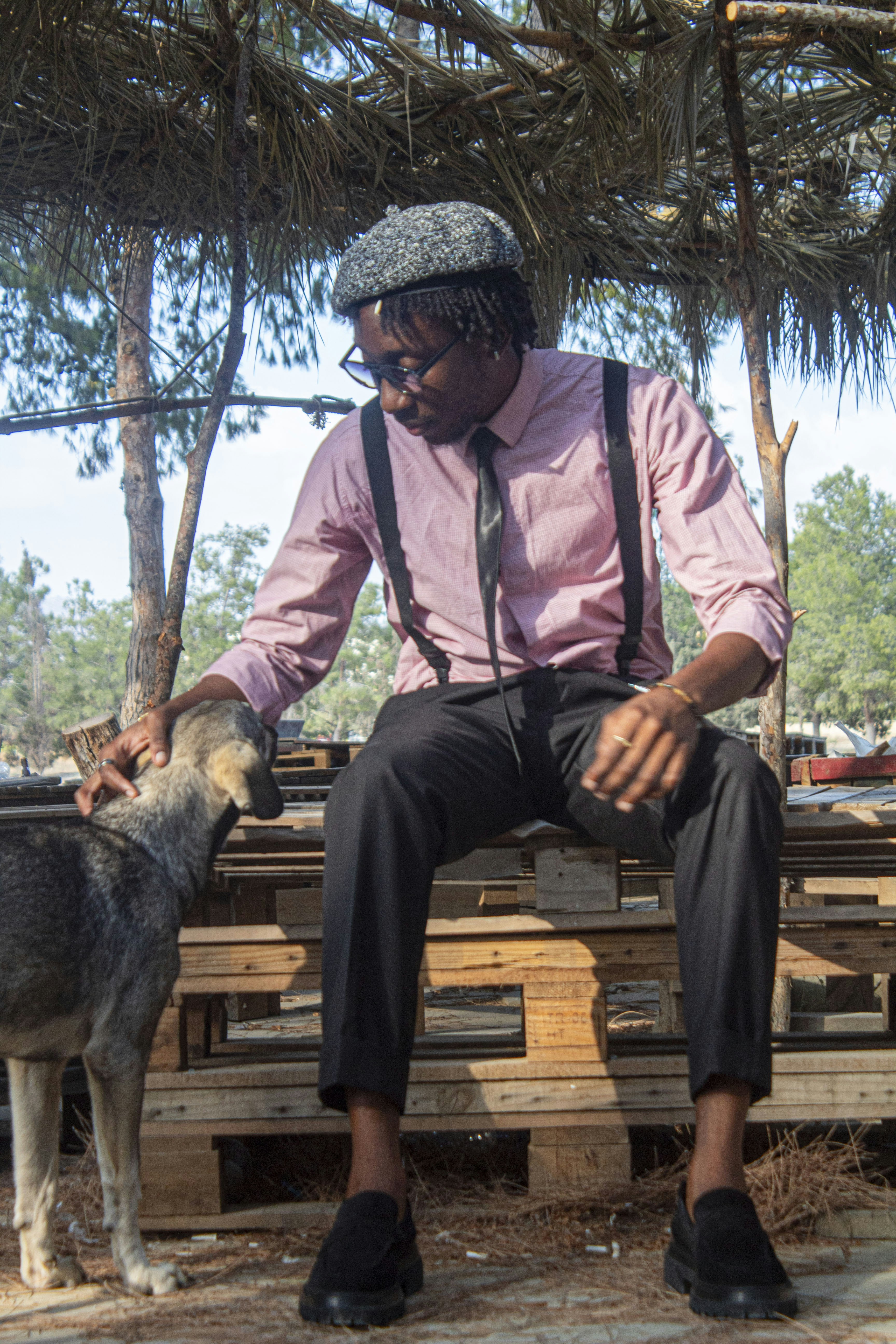 A man in a pink shirt and suspenders interacts tenderly with a dog under a shaded area, surrounded by wooden pallets and greenery.