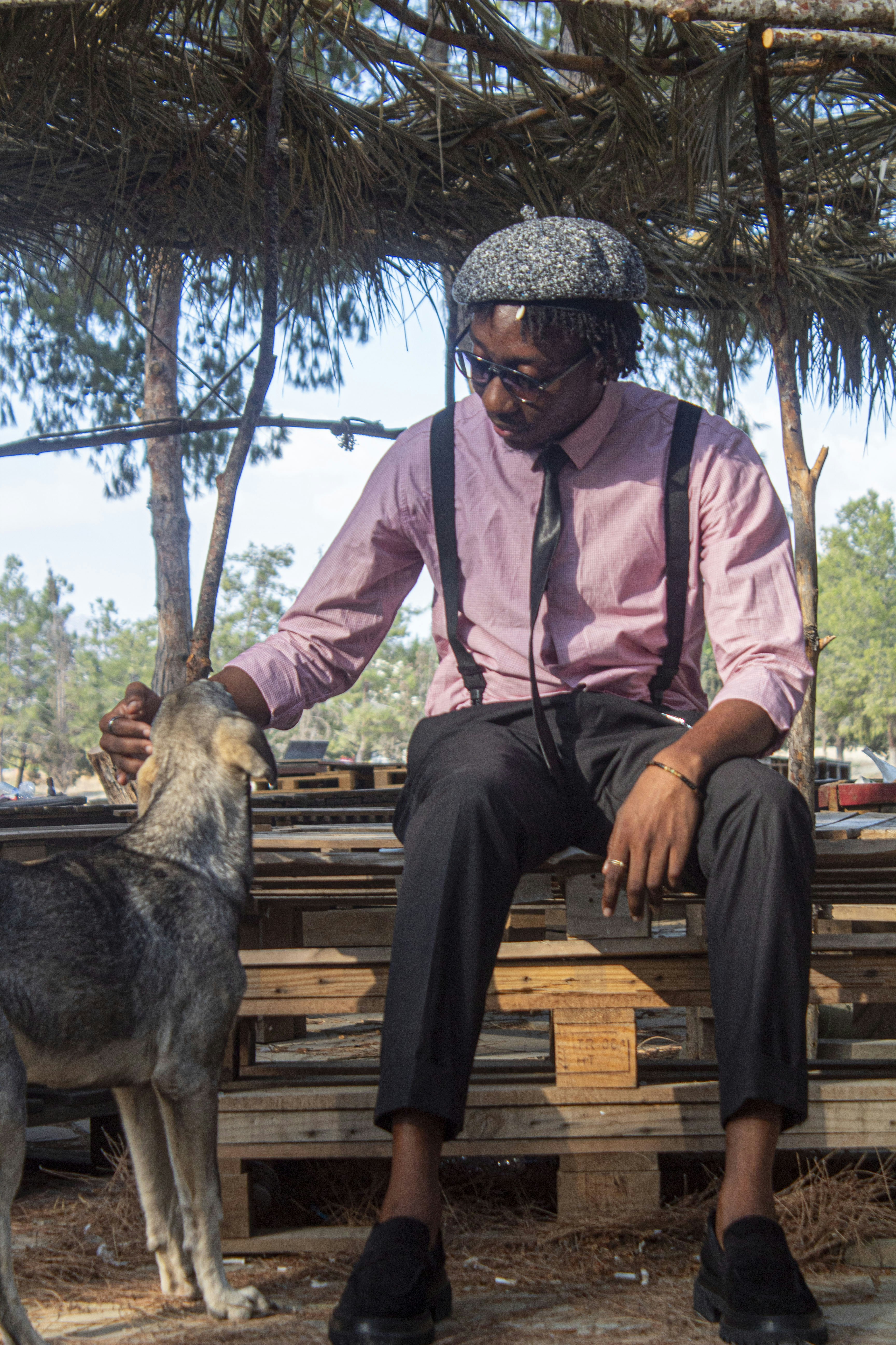 Man in a pink shirt interacts with a dog under a shaded structure, surrounded by wooden pallets and greenery.