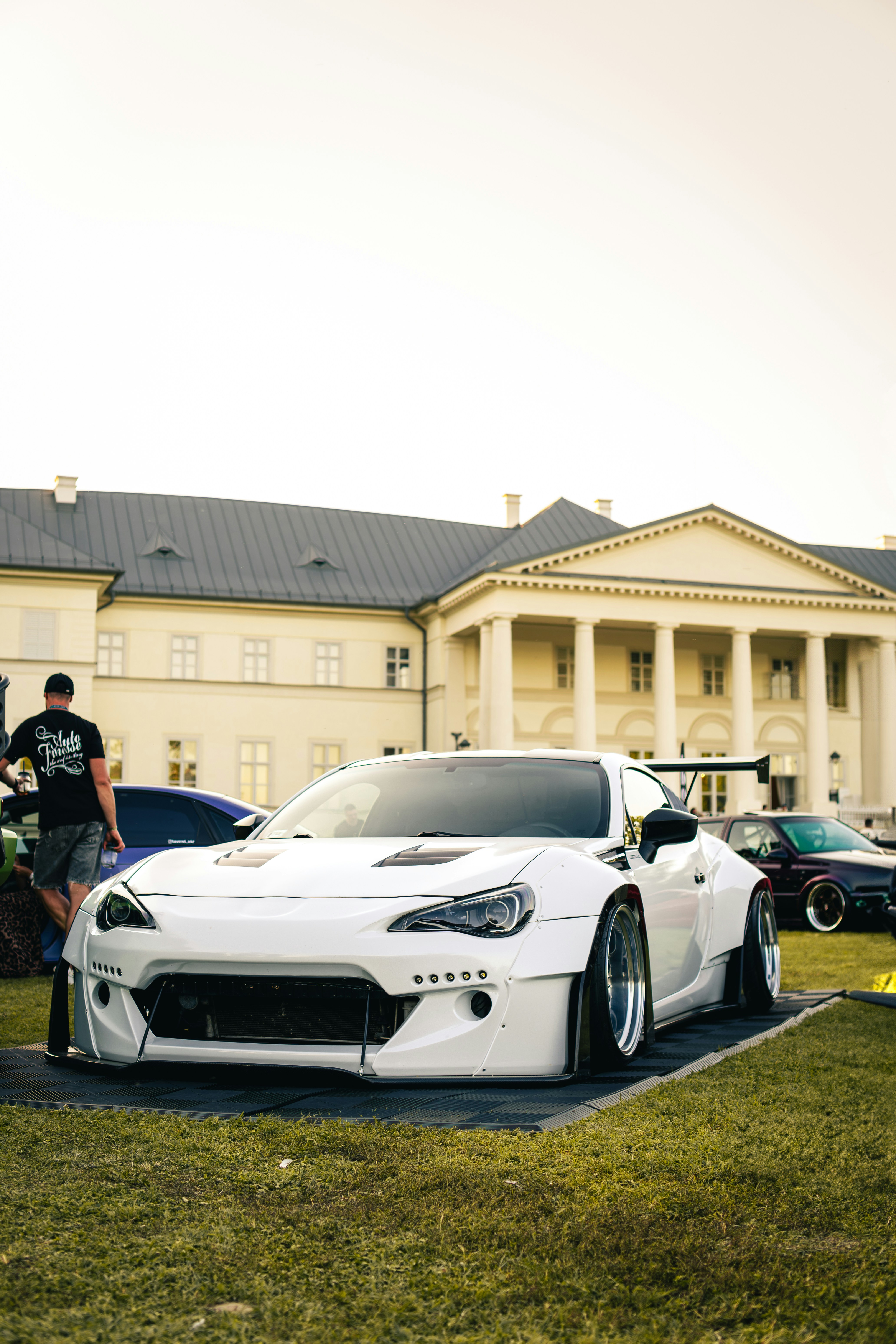 White Widebody Coupe Takes Center Stage at a Car Show Before a Grand Manor