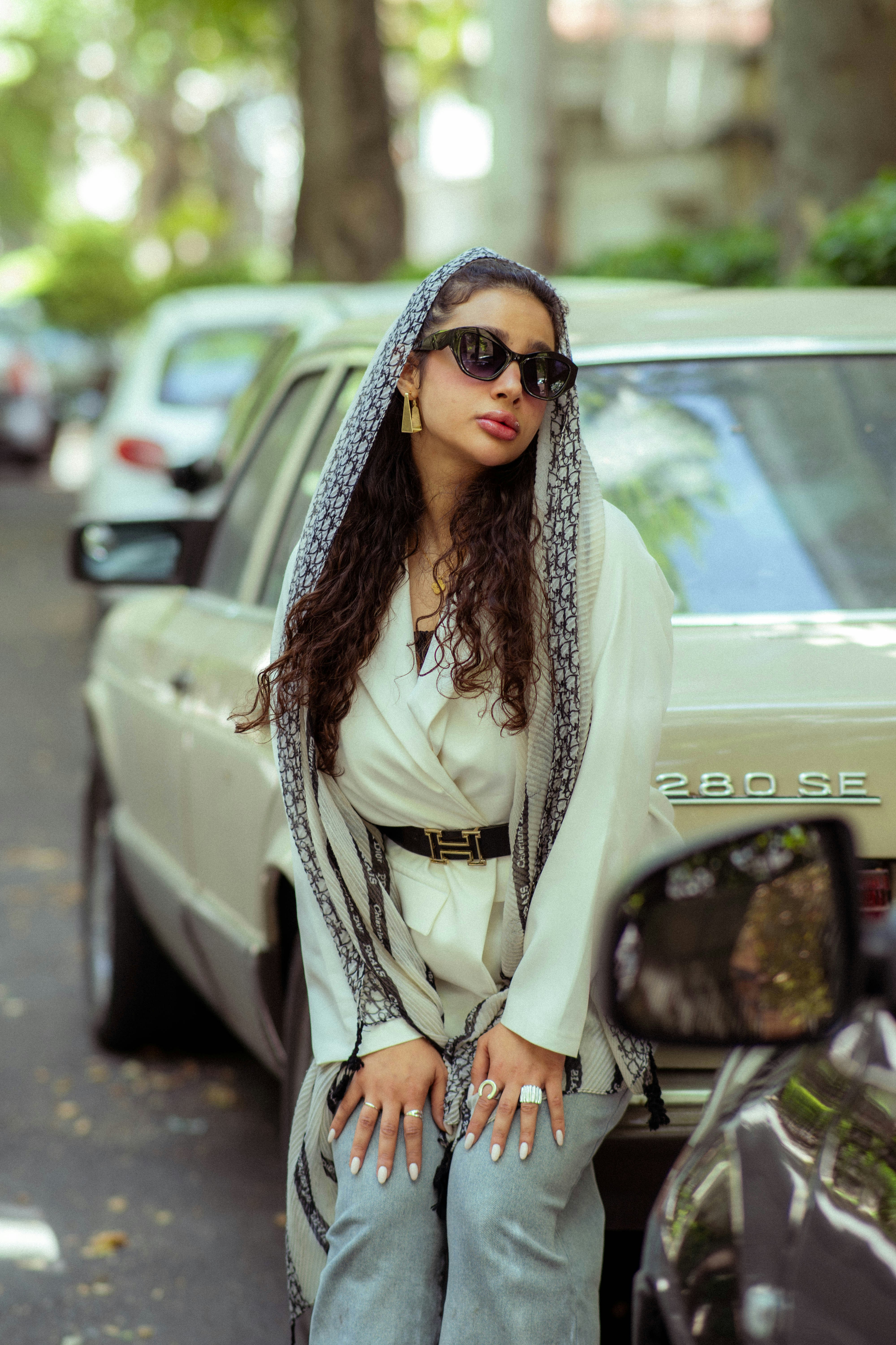 A stylish woman in a flowing outfit and sunglasses poses gracefully beside a classic car, embodying contemporary fashion against an urban backdrop.