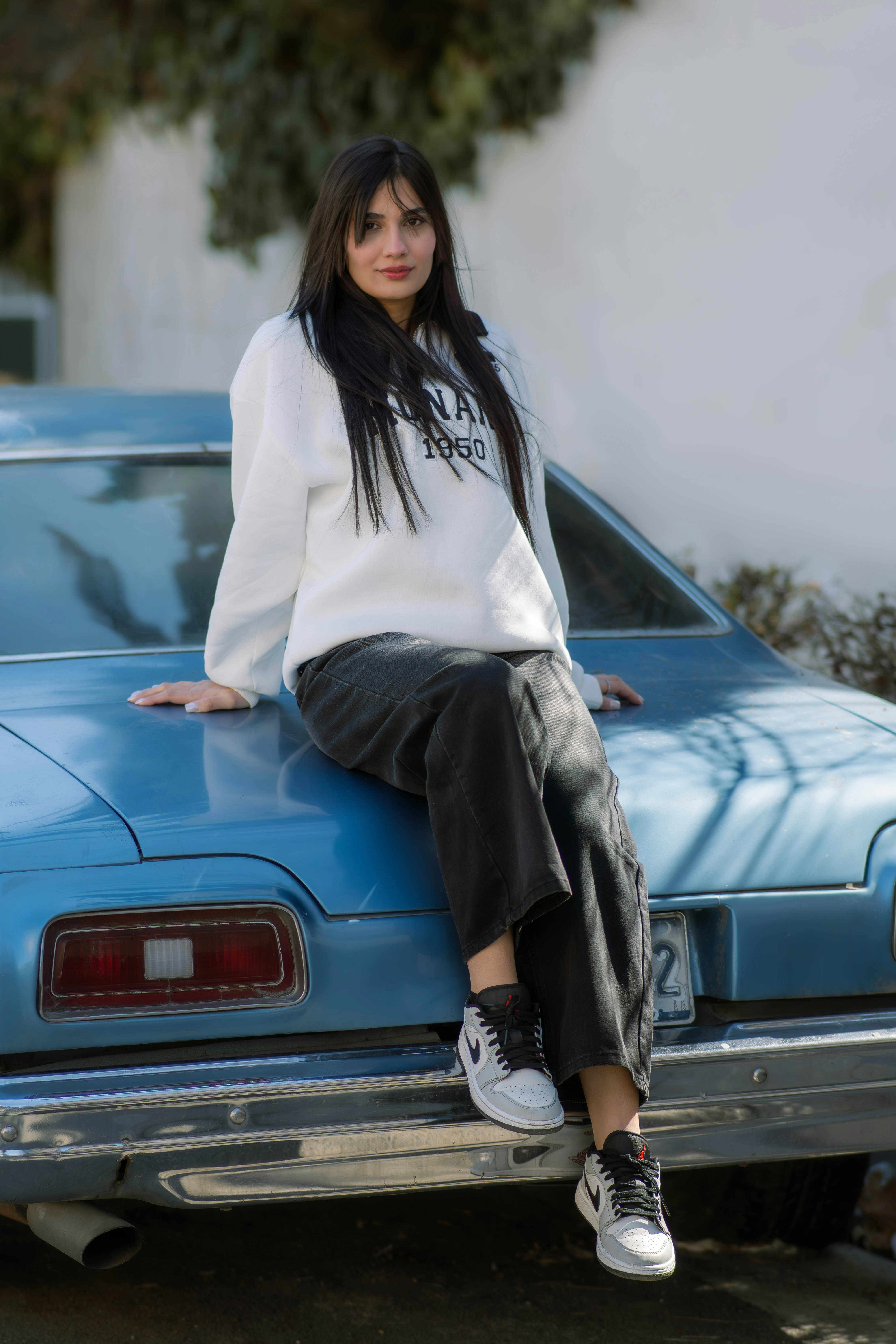 A young woman sits casually on the trunk of a classic blue car, dressed in a white sweatshirt and black pants, exuding a relaxed urban vibe.