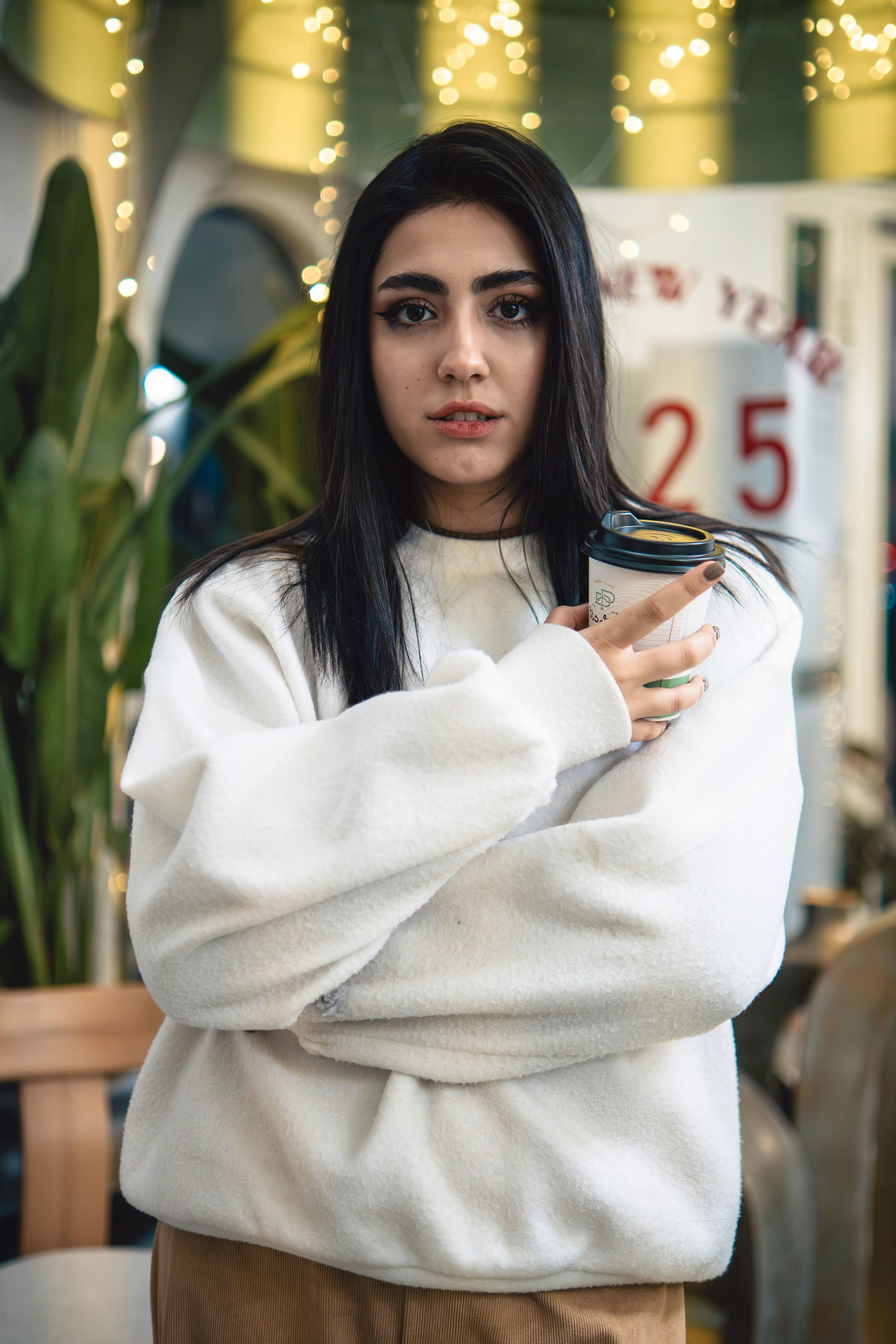 Young woman in a white fleece sweater holding a coffee cup, surrounded by greenery and soft lighting.