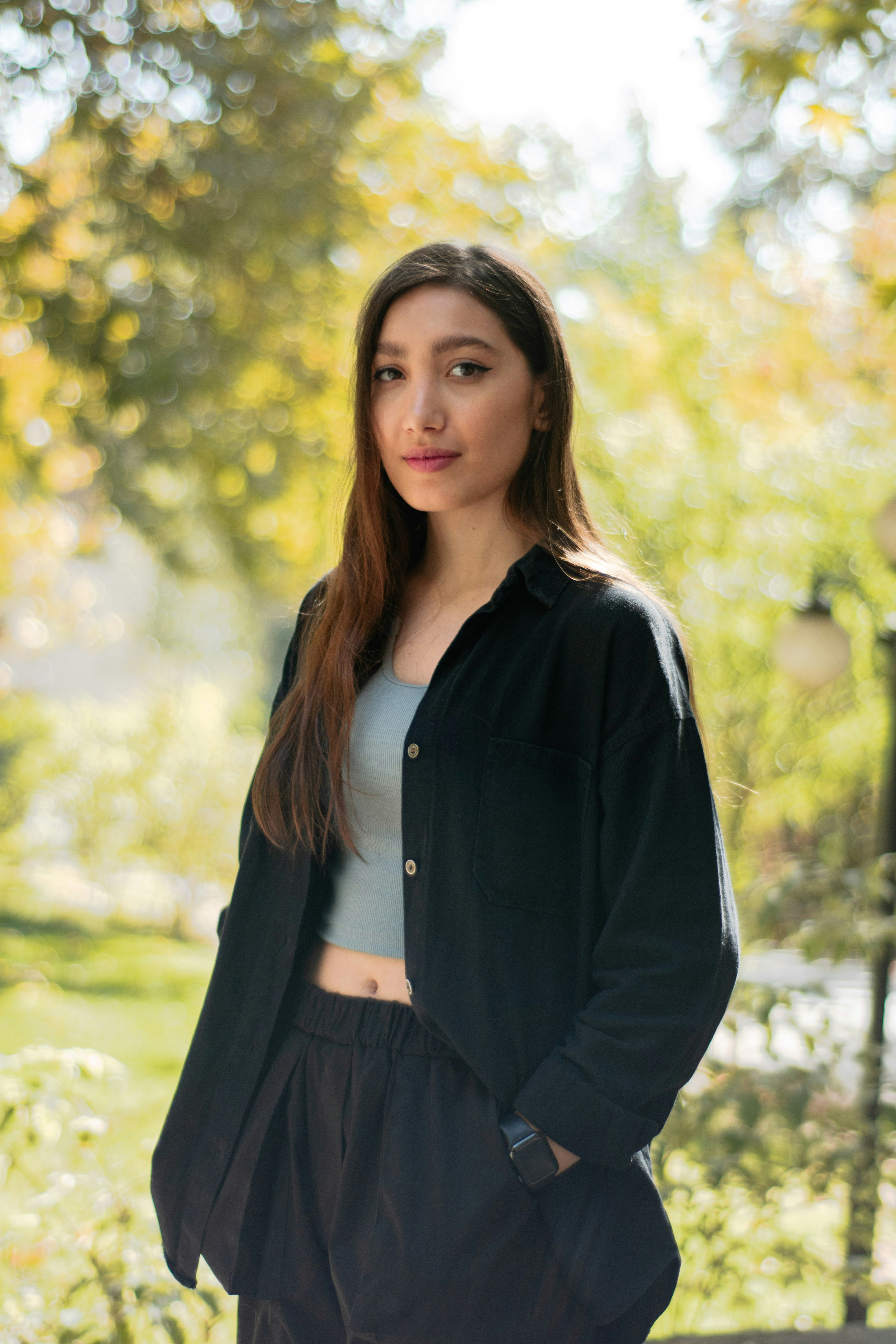 A young woman stands confidently in a sunlit garden, wearing a black shirt and relaxed pants, with soft bokeh in the background. Her serene expression complements the warm tones of the surrounding foliage.