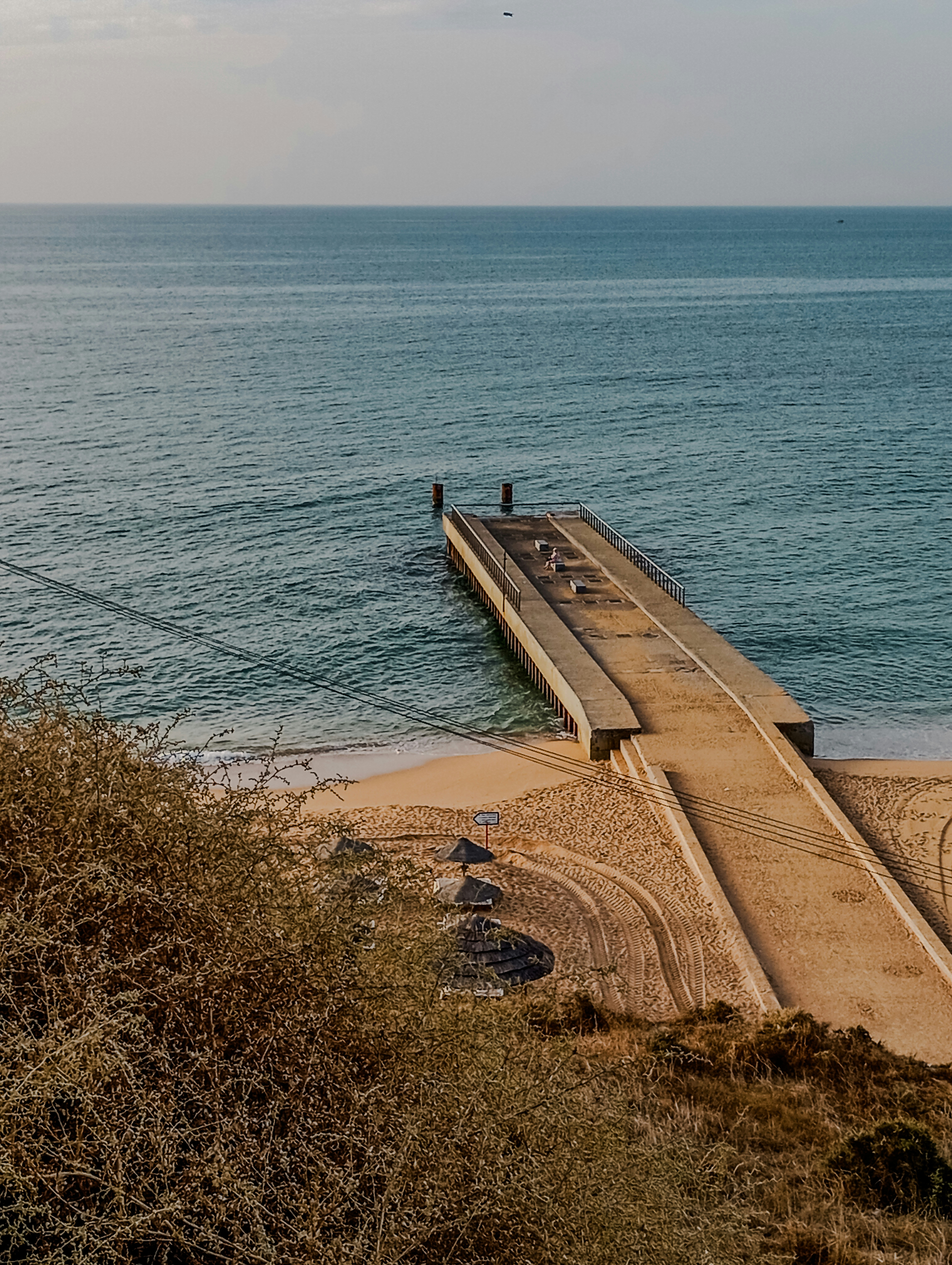 A serene beach scene featuring a wooden pier extending into calm waters, framed by gentle sandy curves and sparse vegetation.