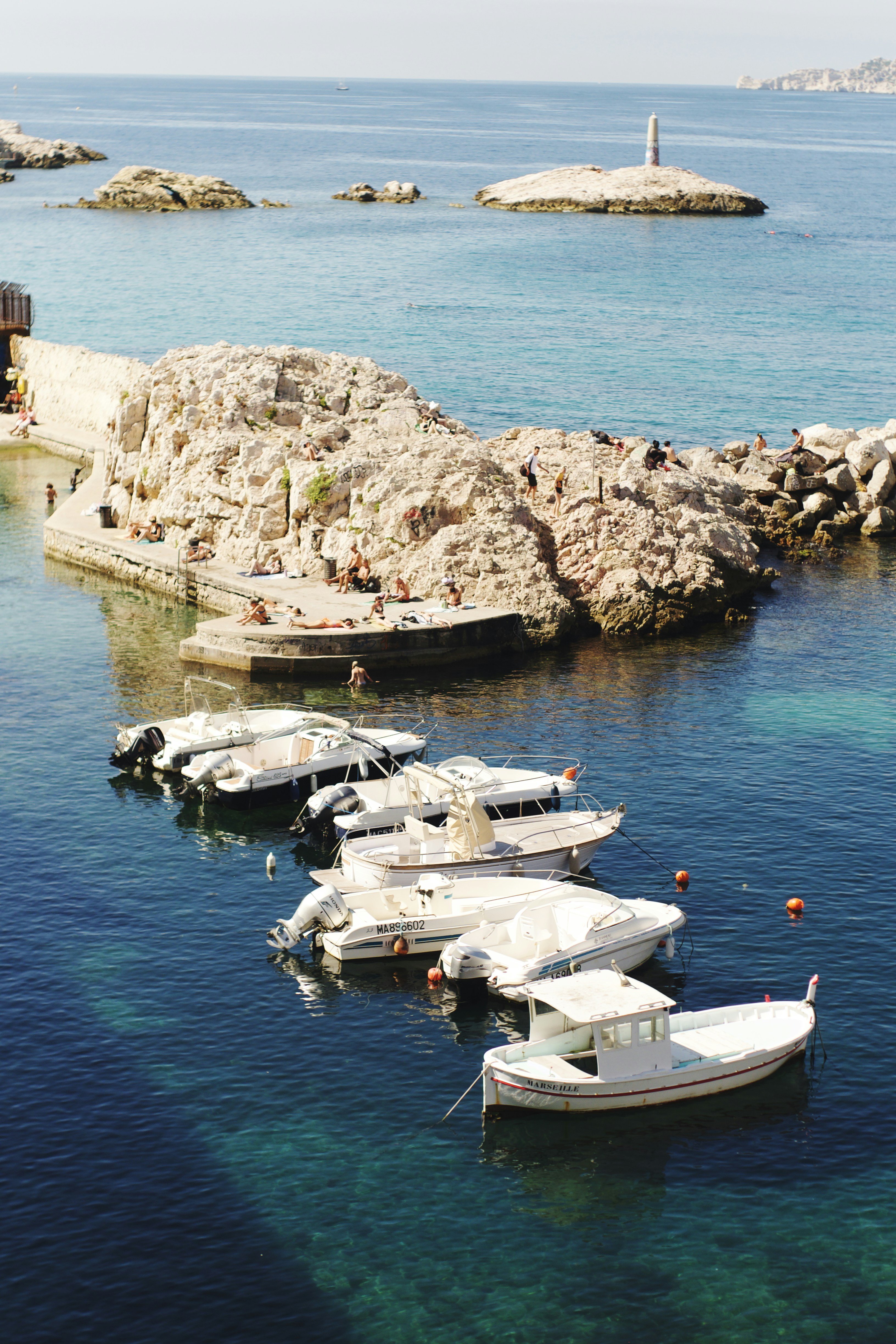 Sunny day at Marseille. Boats floating on the sea. | Several boats moored in clear blue water near rocky shore.