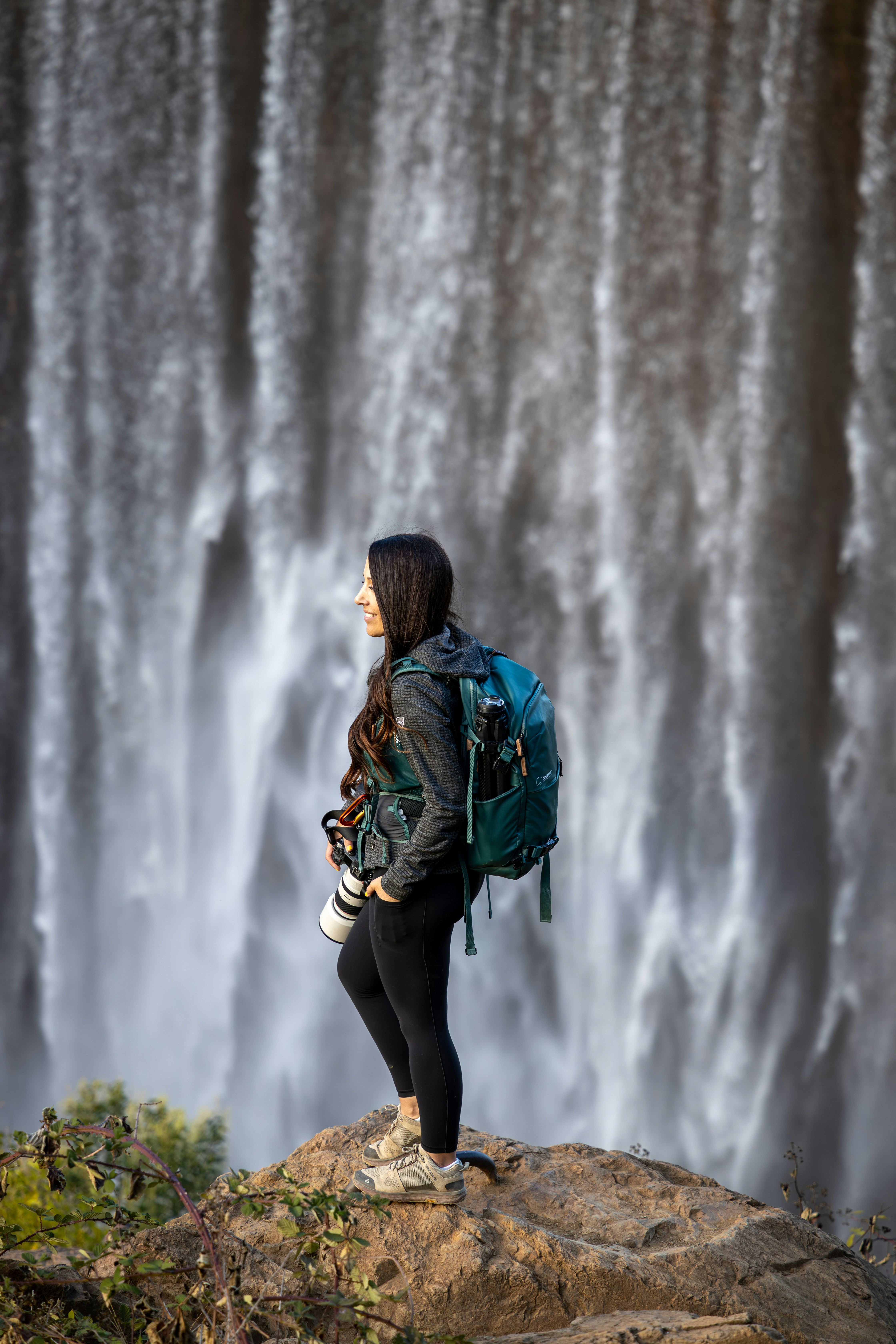 Hiker pausing at the edge of a rocky outcrop, with a majestic waterfall cascading in the background. The scene captures the spirit of adventure and connection with nature.