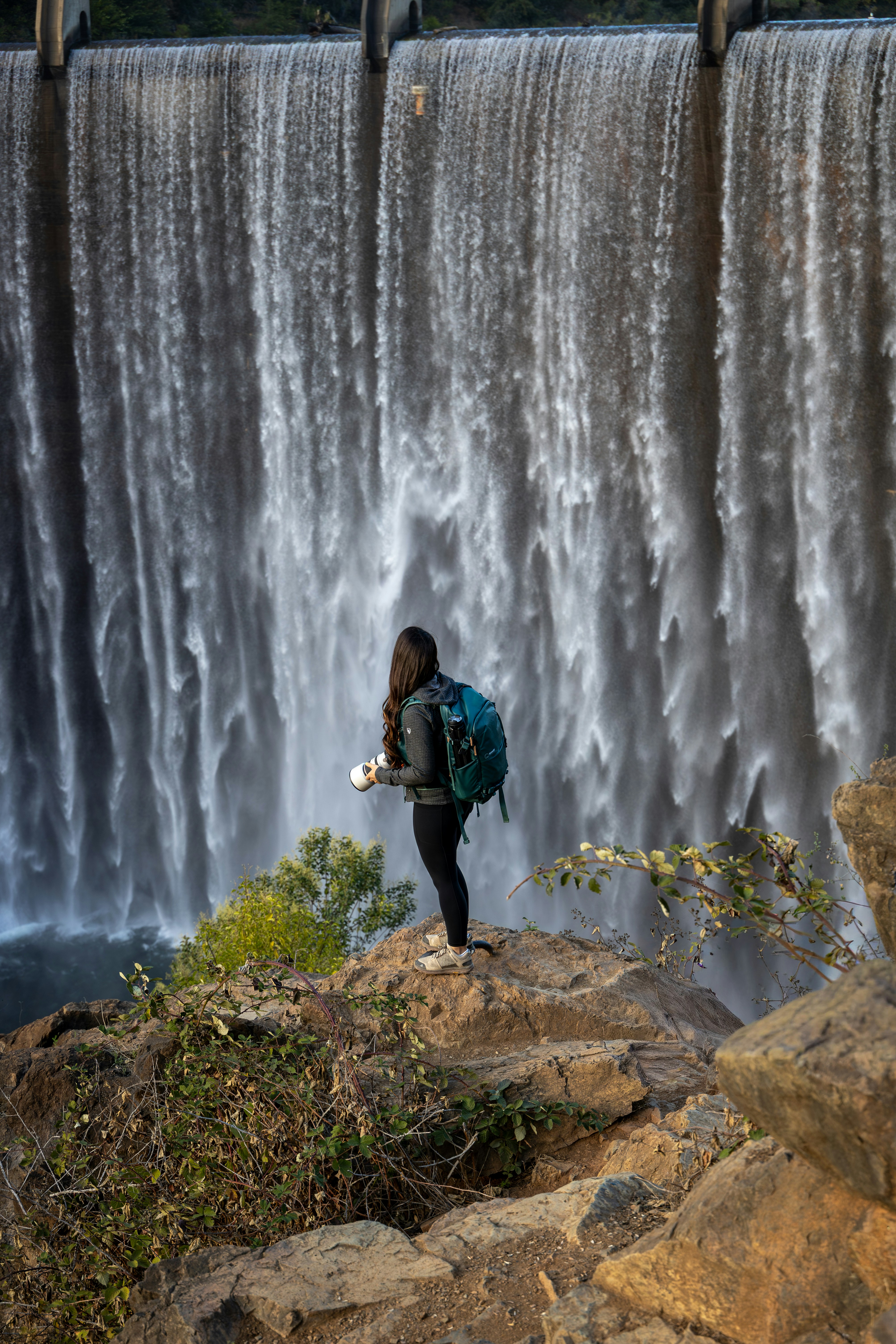 Woman with backpack stands before a large waterfall.